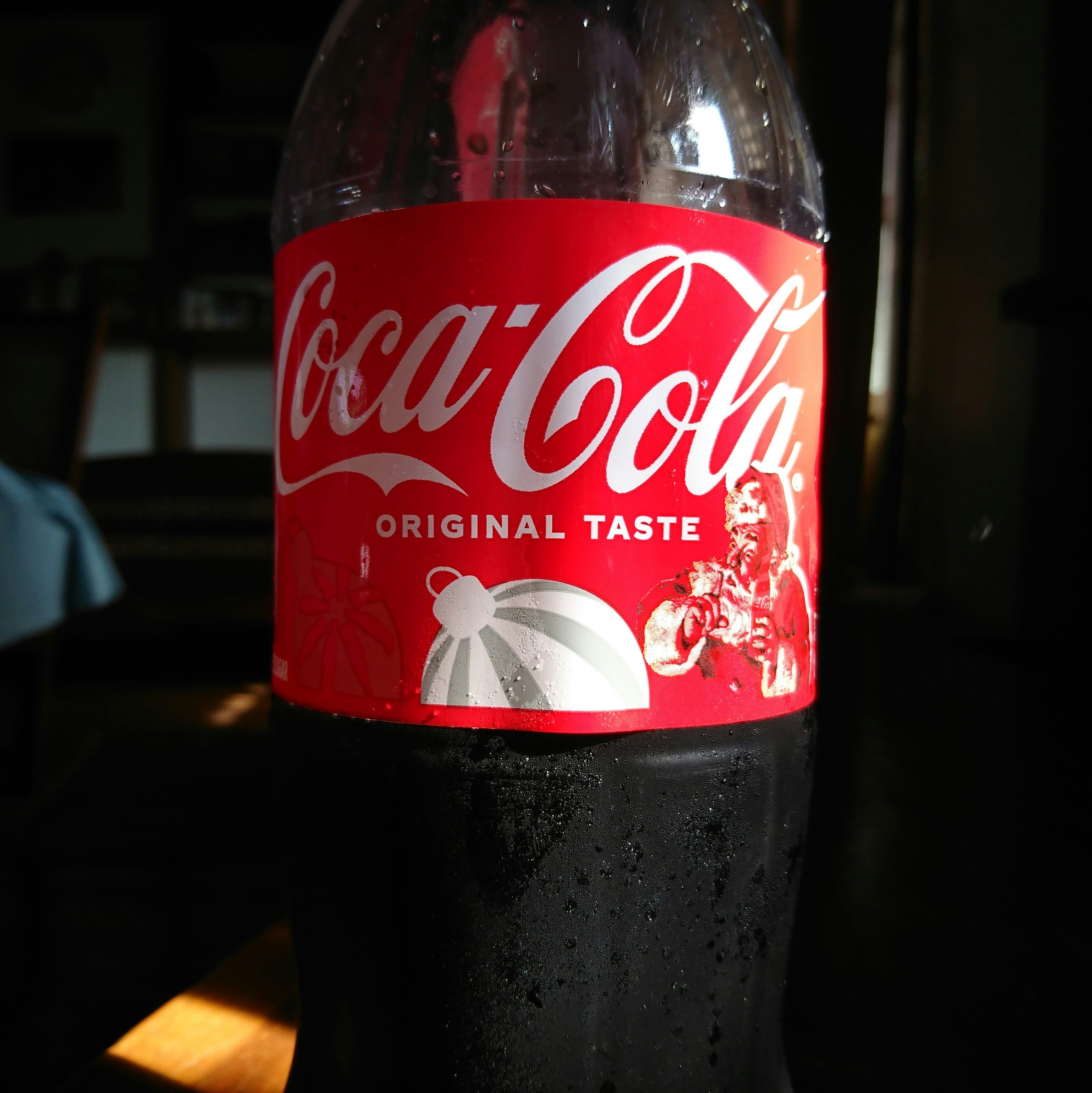 Coca-Cola bottle with iconic red label on a dark surface, illuminated by a warm single light revealing condensation. Close-up photograph highlighting branding and glossy packaging.