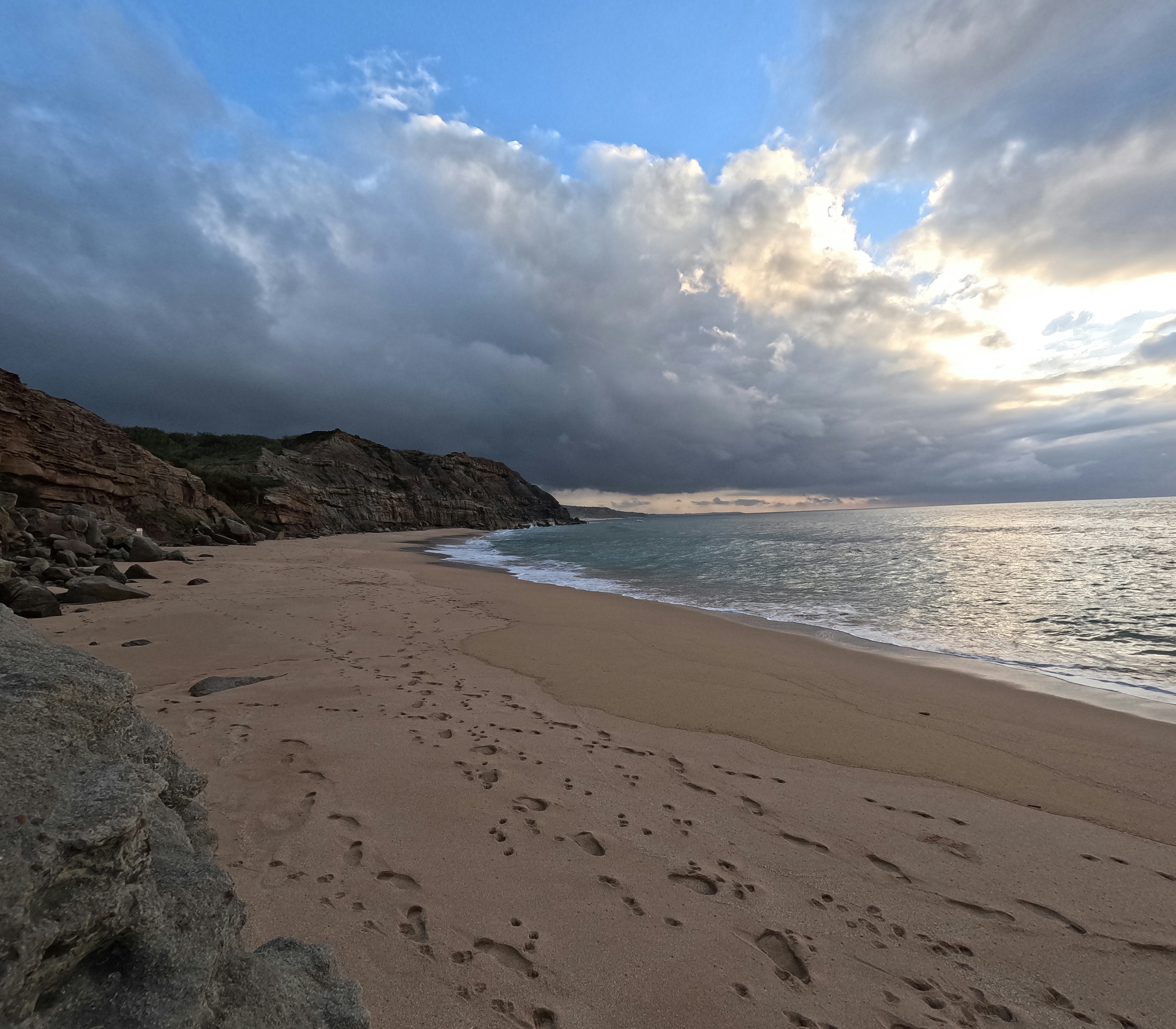 Coastal scene of a sandy beach with a rocky headland on the left, calm sea on the right, and footprints leading toward the water under dramatic clouds.