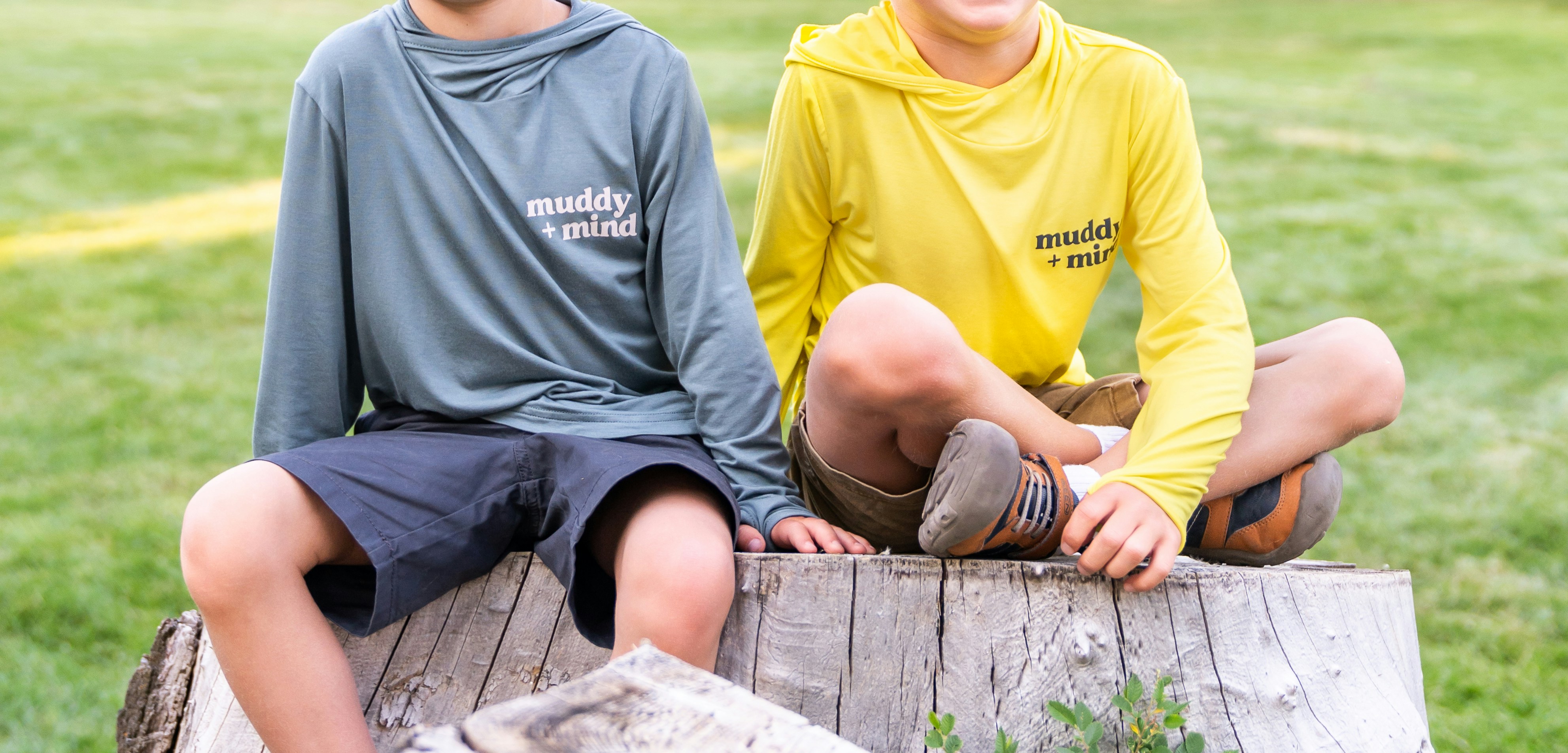 Two boys sitting on a weathered log, one in a yellow shirt and the other in a gray shirt, enjoying a sunny day outdoors.