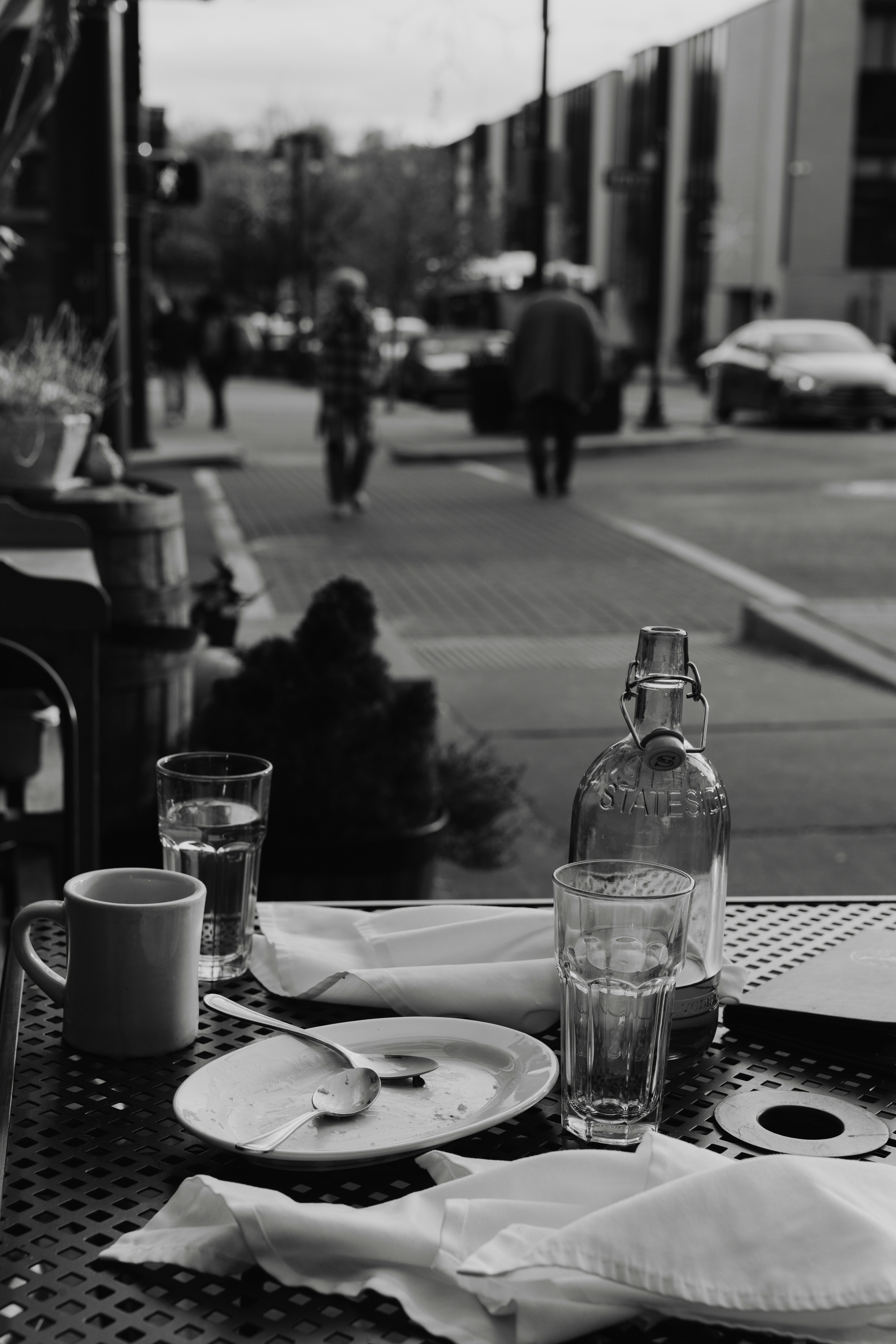 Mono Café Table: Glass Bottle, Plate and Napkins Anchoring a Quiet City Street