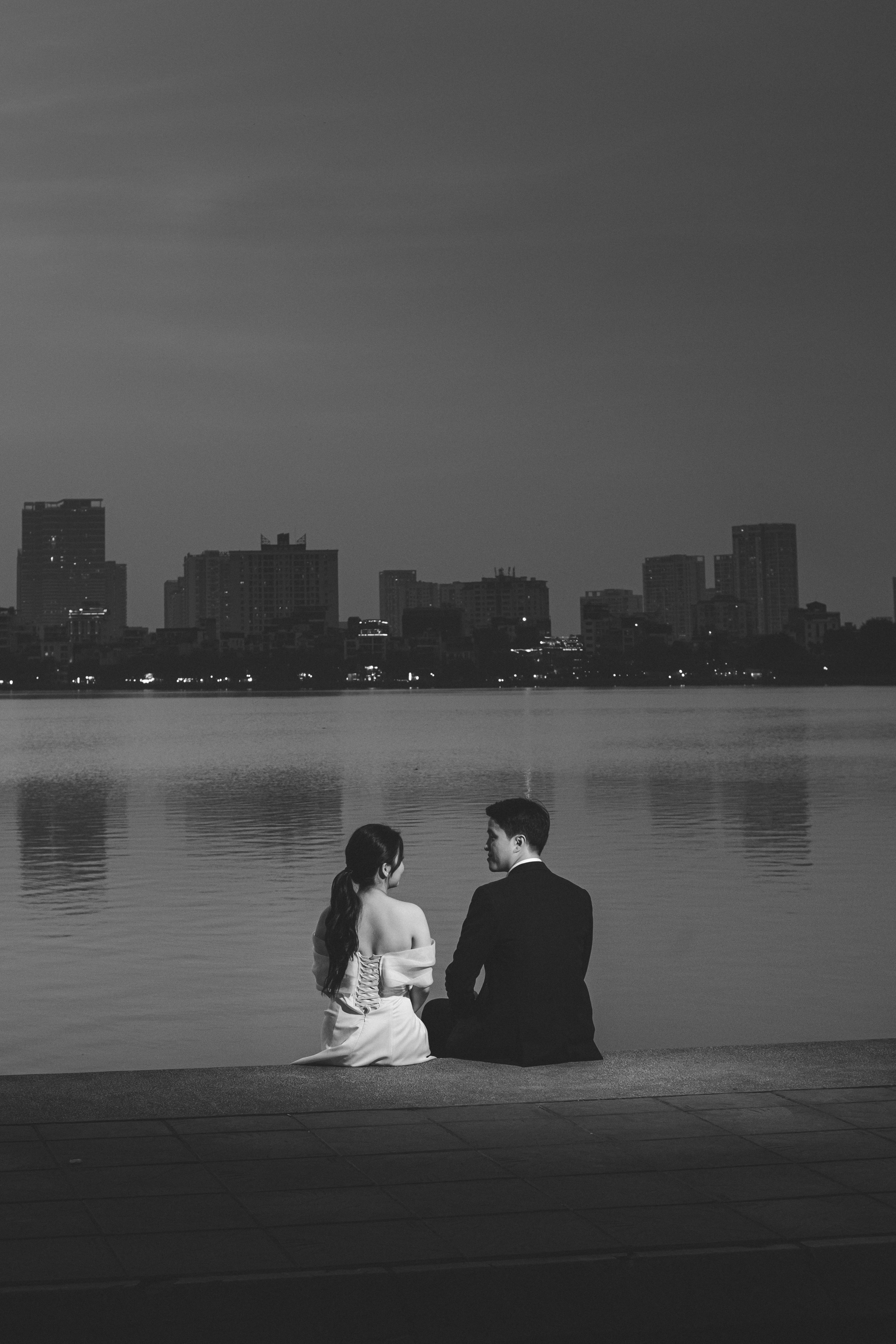 Couple seated by a tranquil lake, silhouetted against a city skyline at dusk. The scene captures a serene moment of connection.