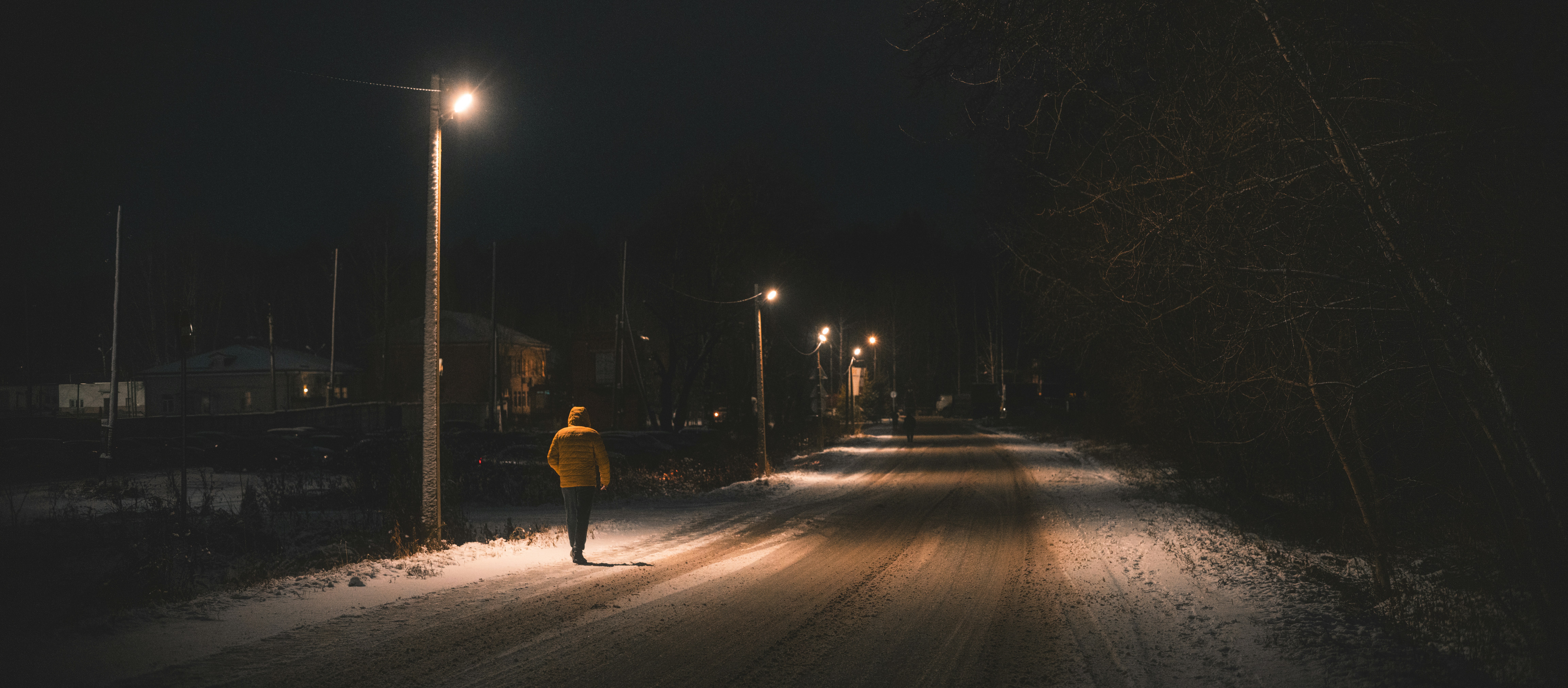 A lone figure in a yellow coat walks down a snow-covered road illuminated by streetlights at night.