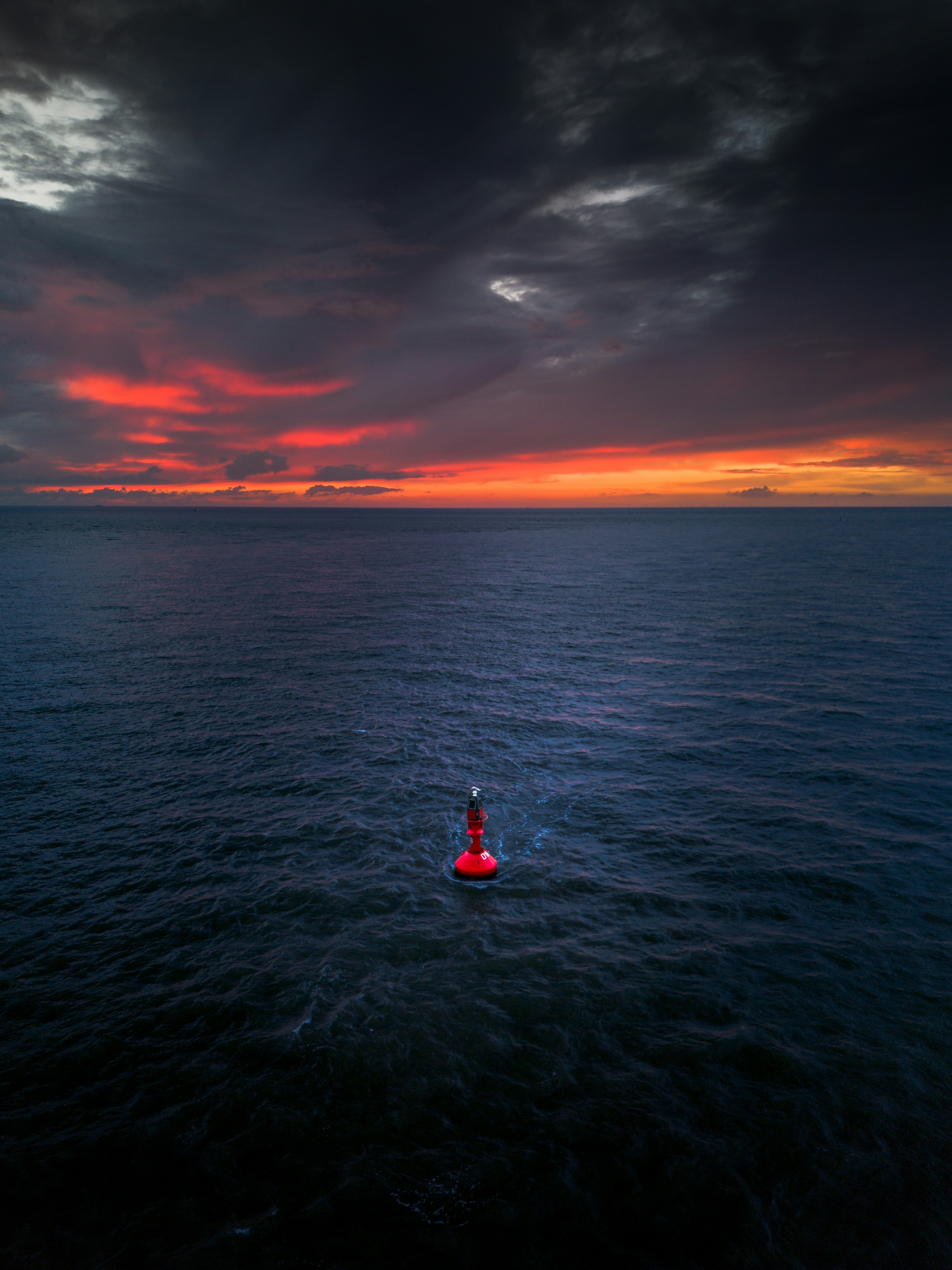 Red buoy anchors the foreground as dark, rippling water leads to a horizon lit by orange and red sunset. Drone photograph captures the buoy against a vast sea and a dramatic, cloud-filled sky.