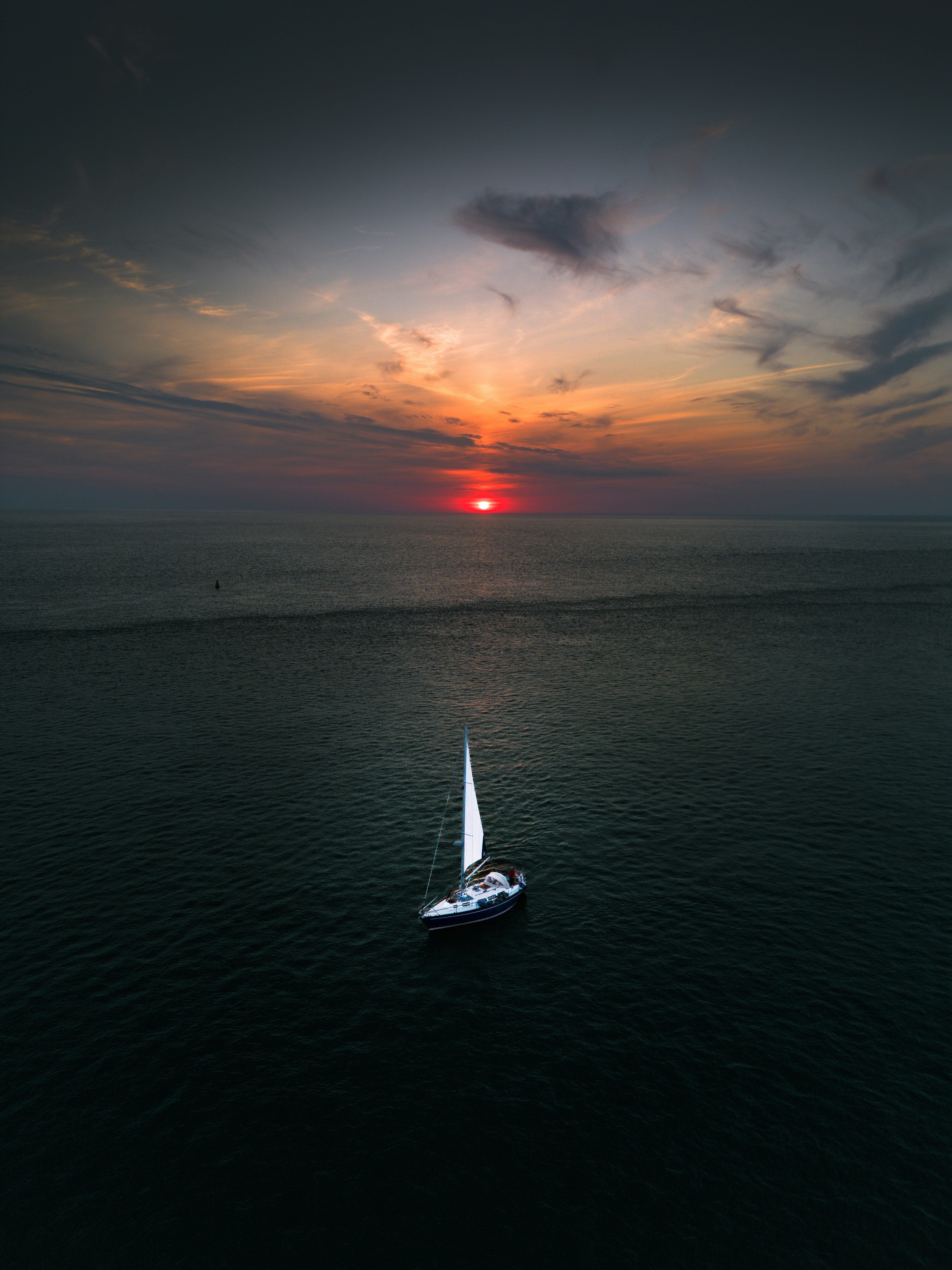 Sailboat glides across a calm sea toward a crimson sunset, with wispy clouds drifting overhead.