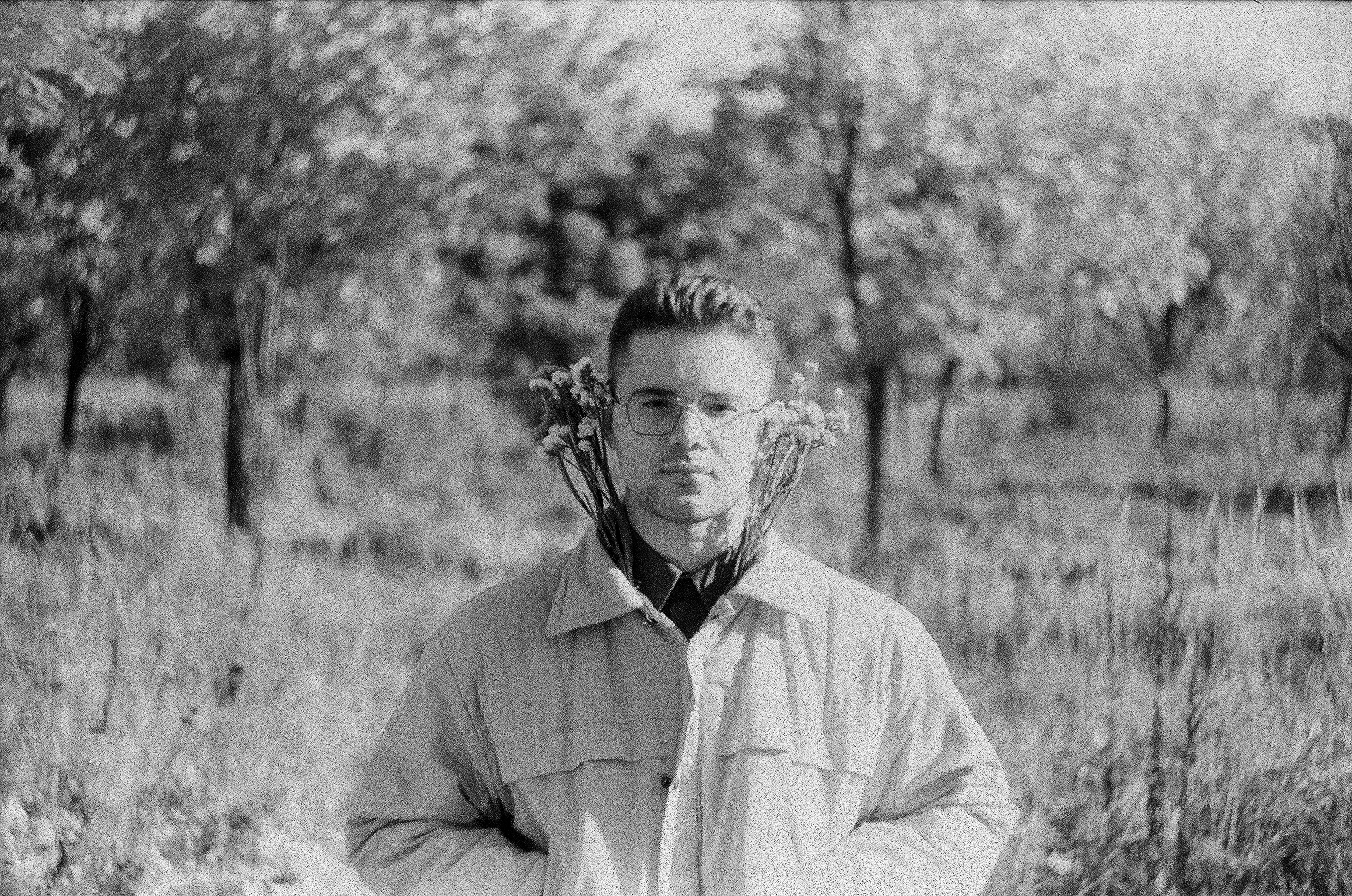 Black-and-white photograph of a man in a light jacket standing in a sunlit field, with a bouquet of wildflowers tucked behind his neck. The composition centers the subject against a softly blurred backdrop of trees and grass.