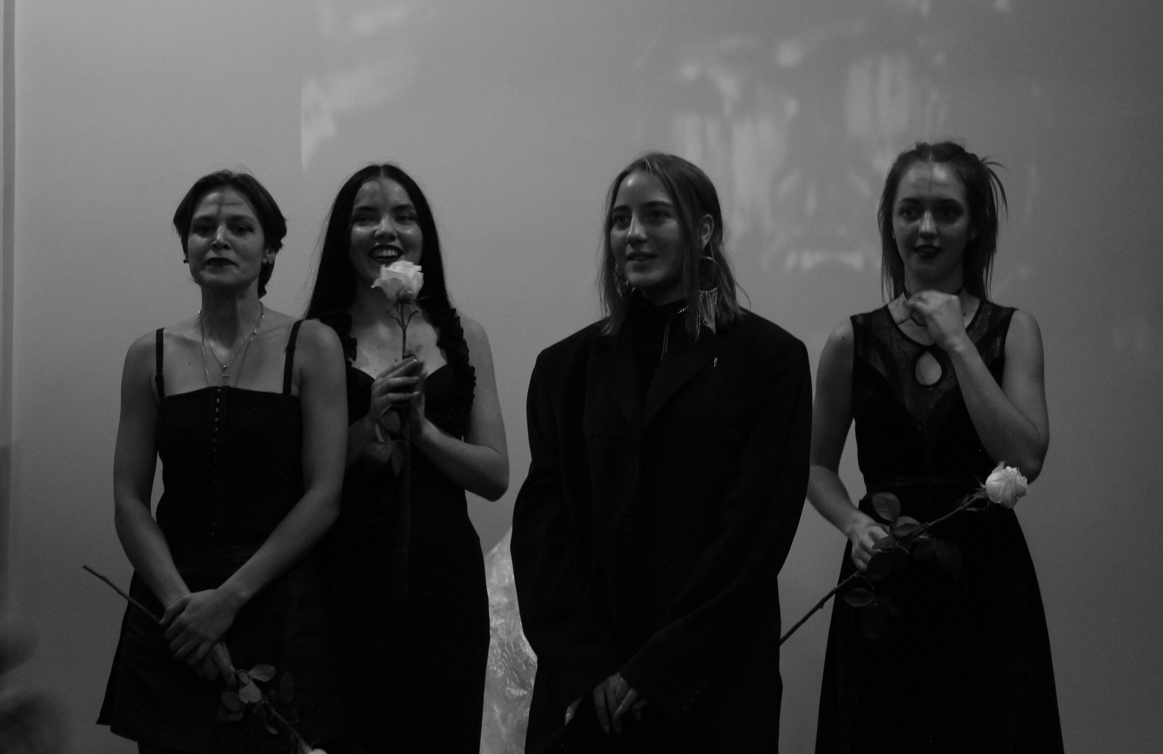 Four women dressed in black attire, each holding a white rose, convey a sense of unity and elegance against a softly blurred background.