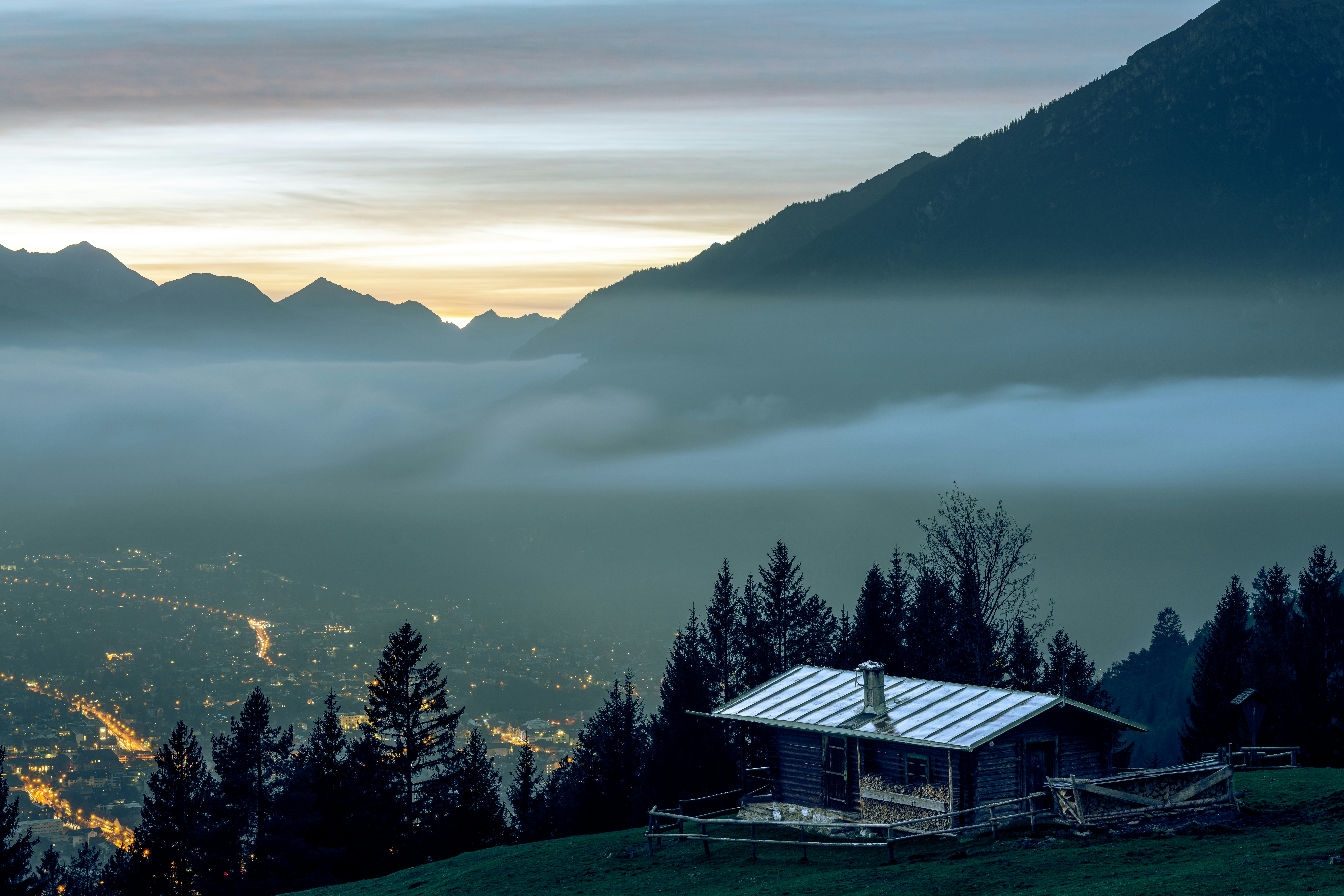 A rustic hut overlooks a misty valley with faint town lights beneath a serene mountain backdrop at dawn.