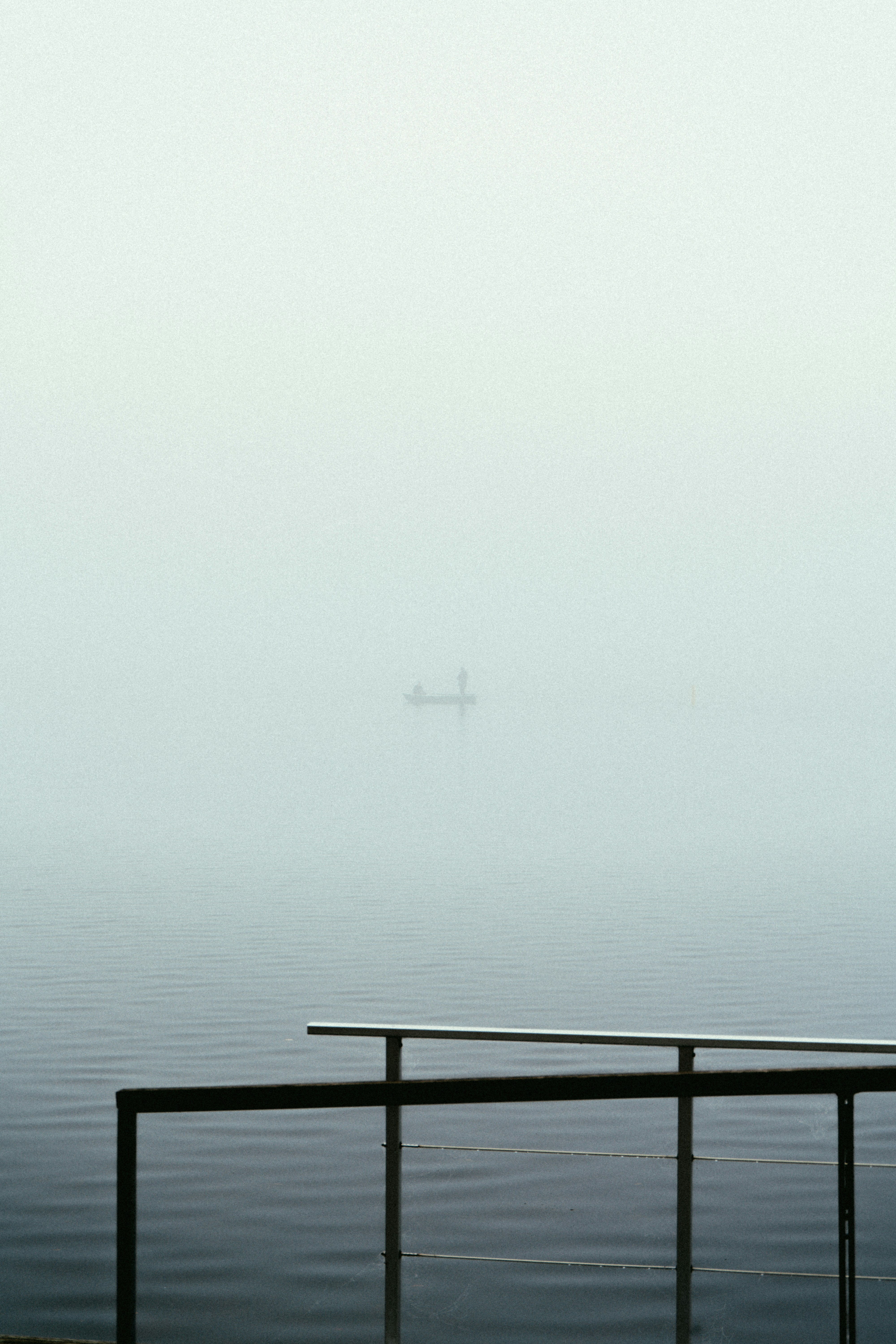 A lone fisherman in a small boat navigates through a dense fog over a calm lake, creating an atmosphere of solitude and stillness.