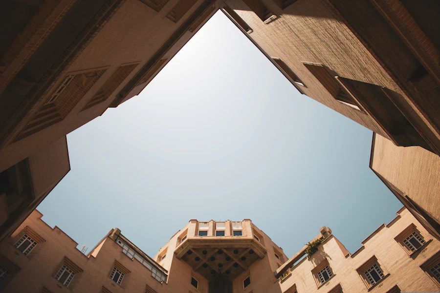 Looking up from a courtyard surrounded by tan apartment buildings, their facades creating a geometric frame around a pale blue sky. A central balcony anchors the composition at the bottom.
