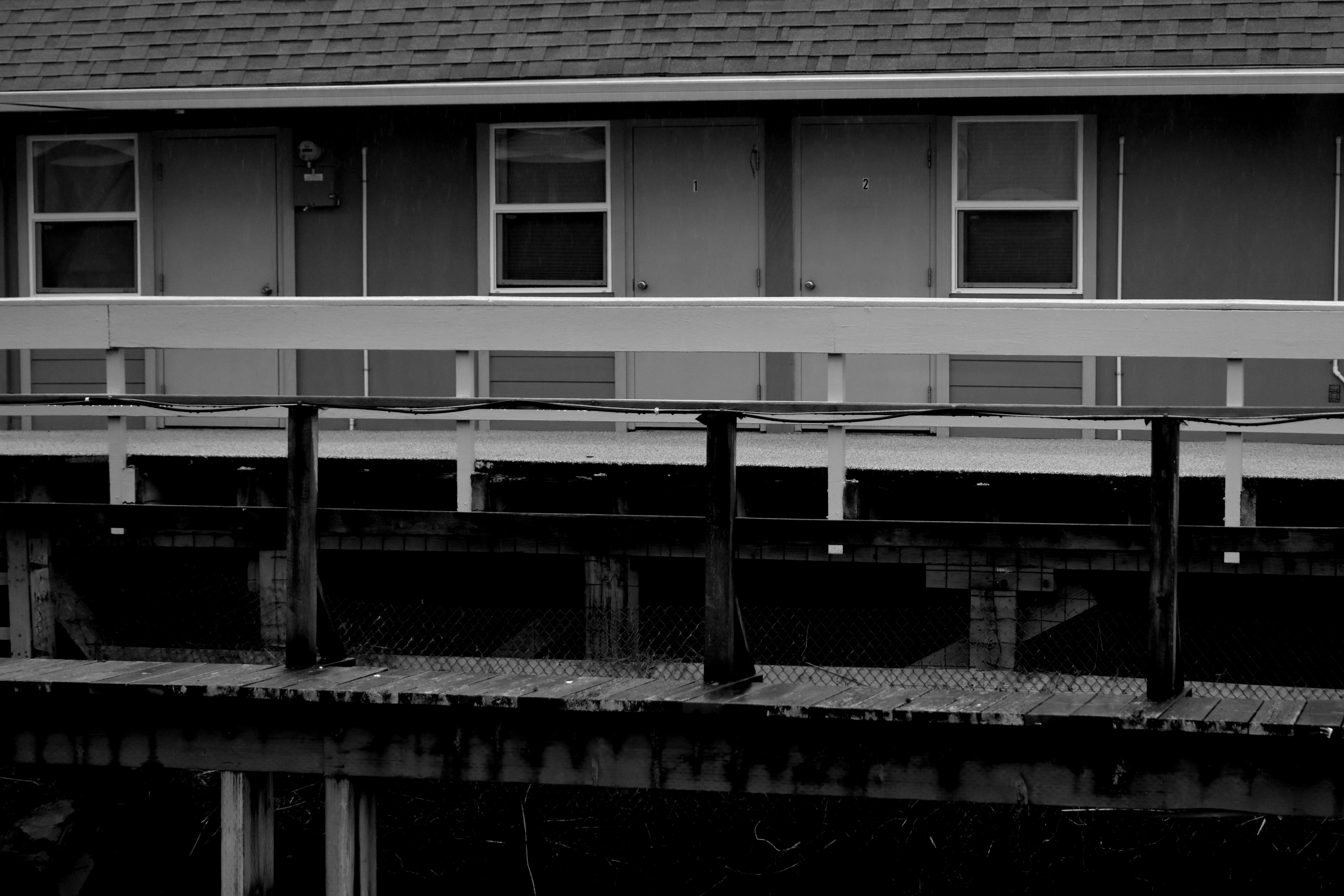 Black and white image of a motel exterior with multiple doors and windows, featuring a wooden railing in the foreground.