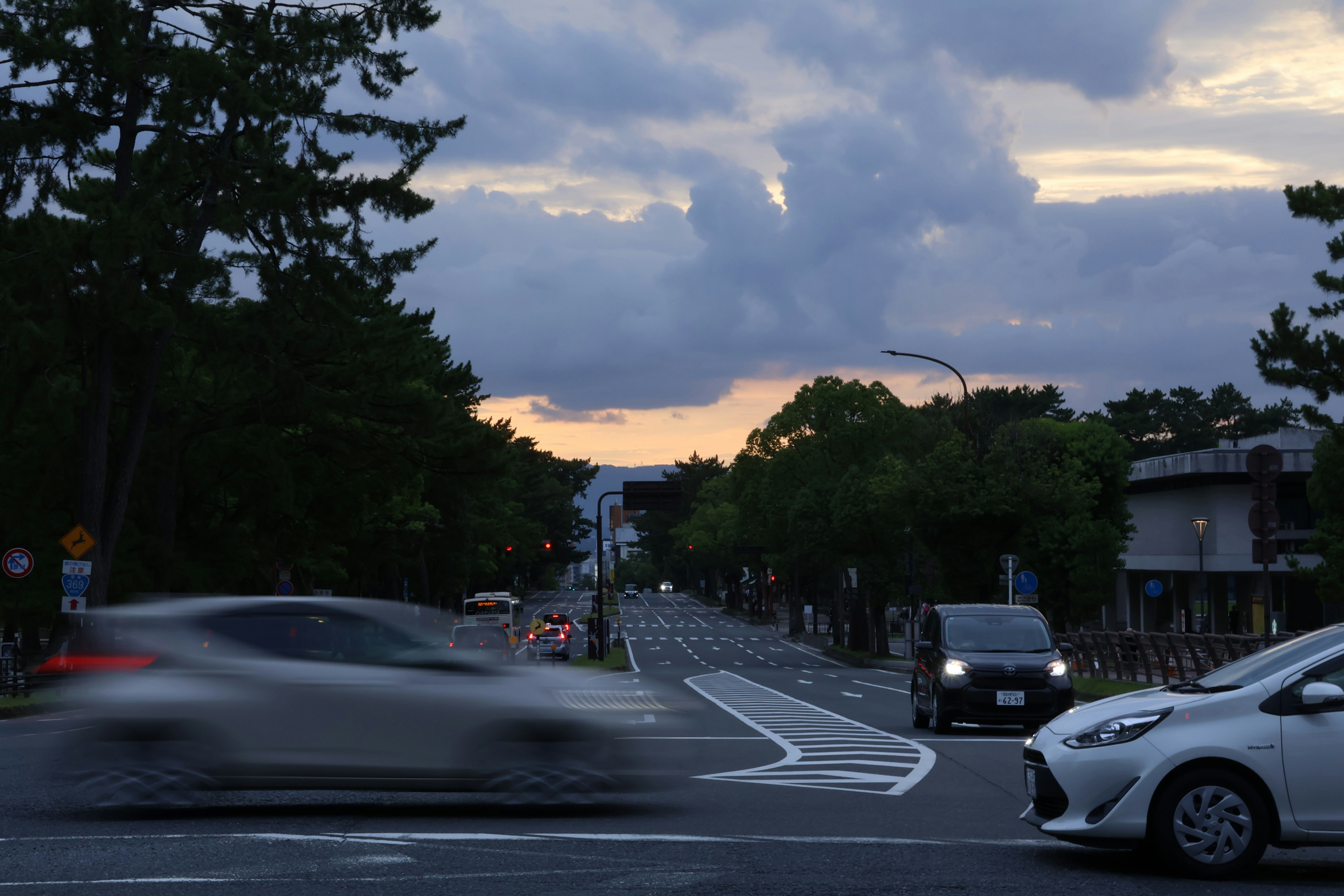 A busy urban intersection at twilight, showcasing blurred motion of vehicles against a serene backdrop of trees and a colorful sky.