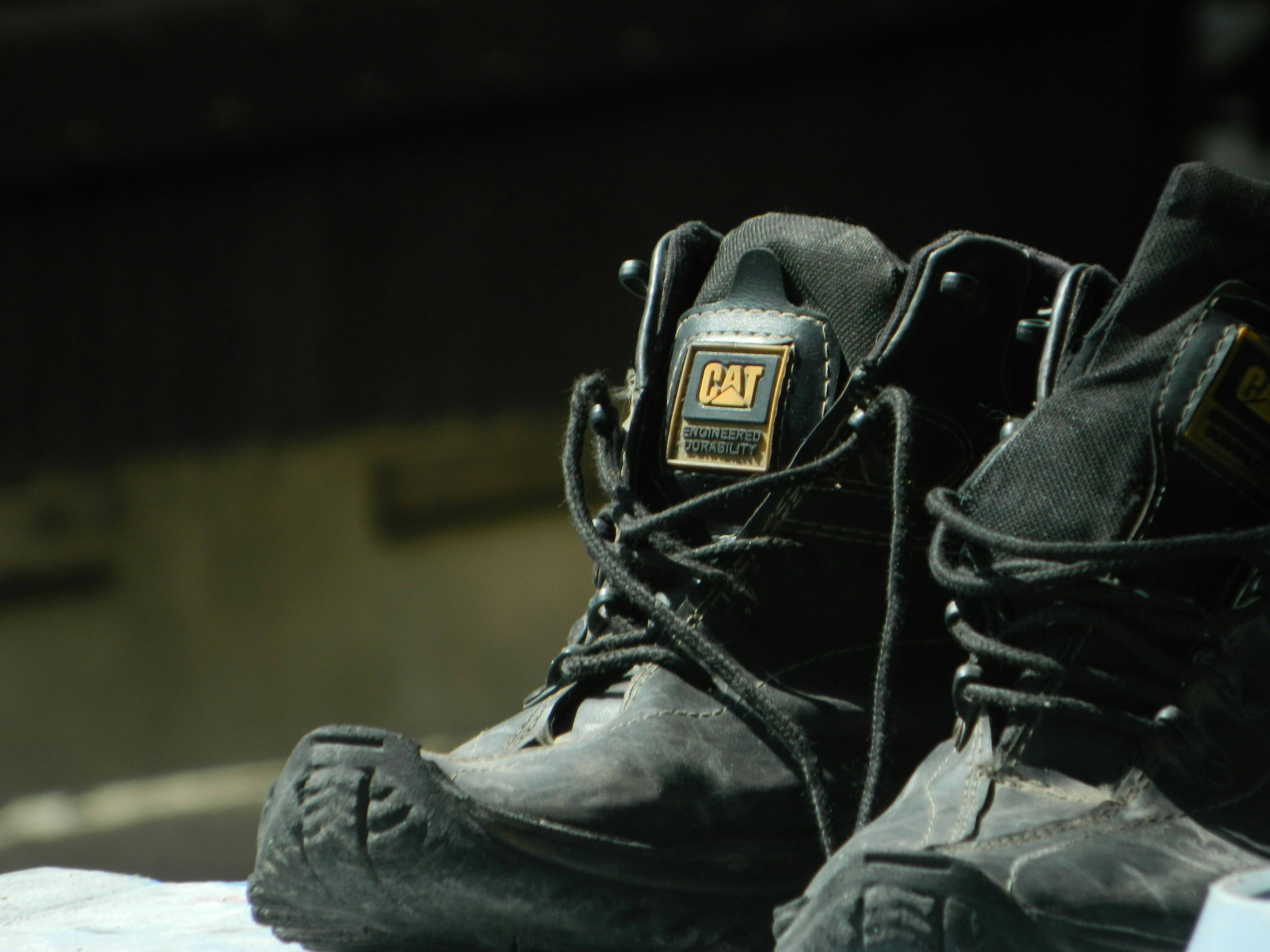 Close-up of rugged CAT work boots with laces and stitching, logo patch on the tongue. They rest on a dim workshop floor, highlighting texture and sturdy construction.