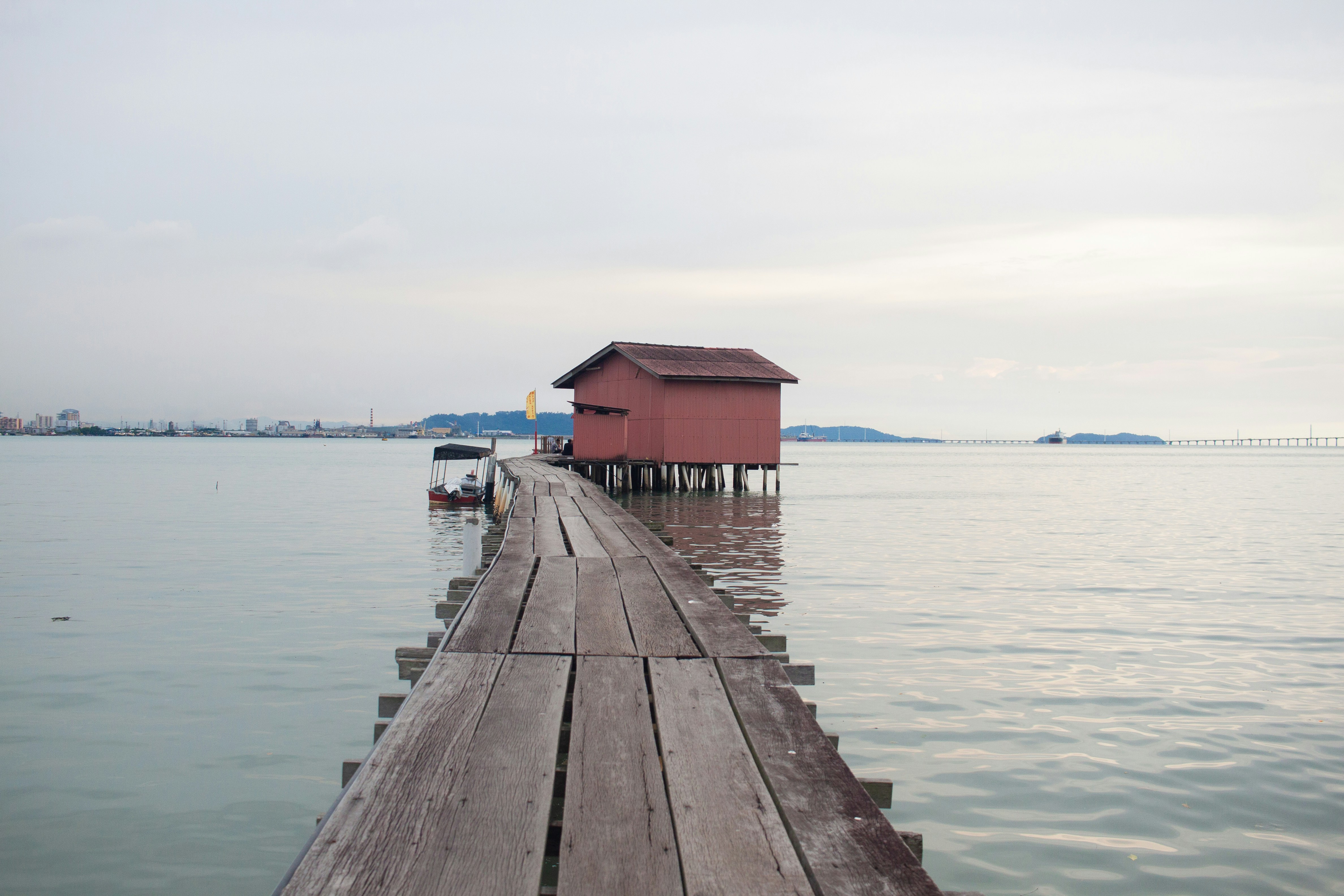 Wooden pier leading to a red stilt house over calm sea under an overcast sky.