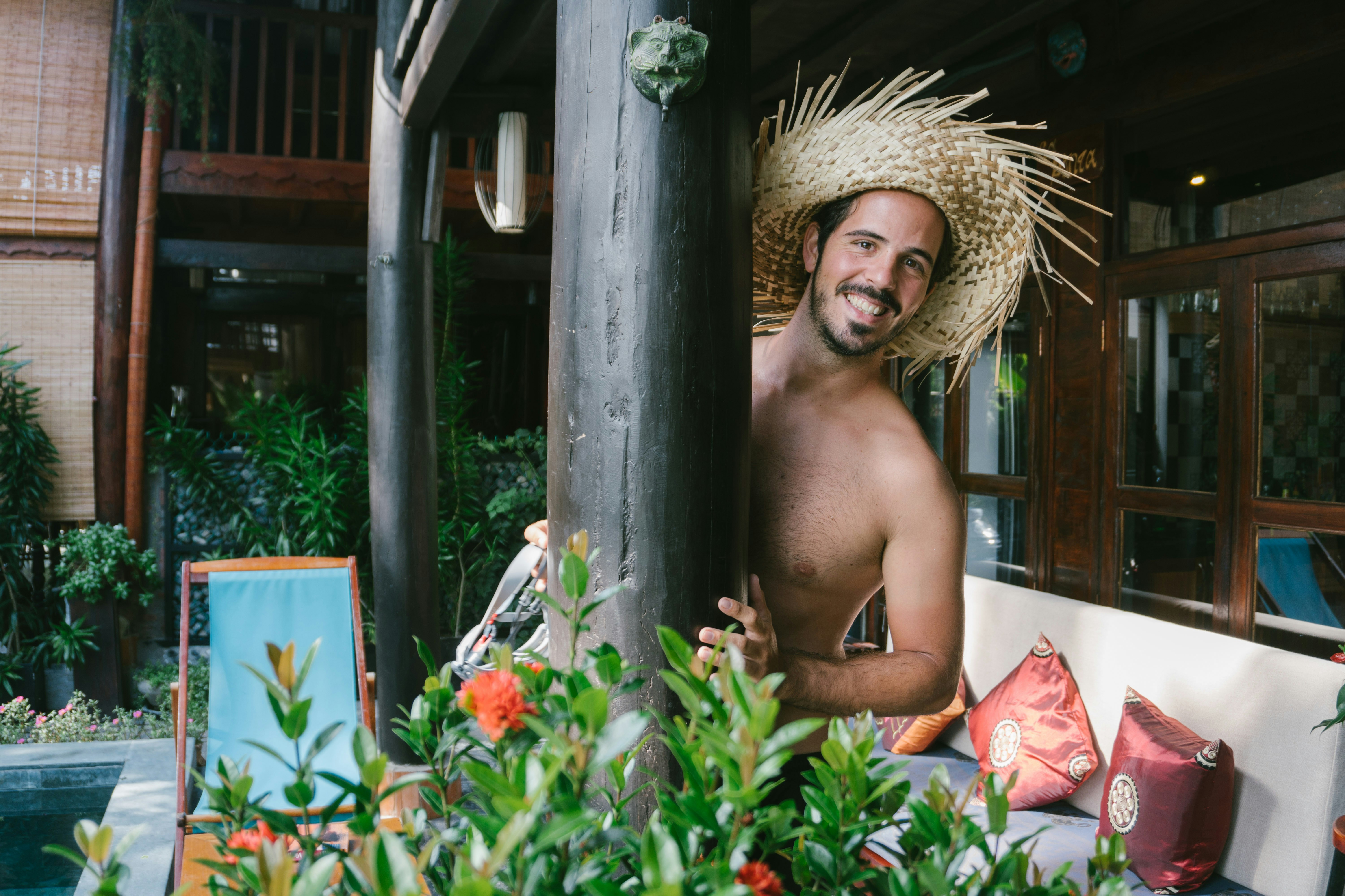 A man wearing a straw hat peeks from behind a wooden post, surrounded by vibrant foliage and cozy seating. The setting exudes a relaxed, tropical ambiance.