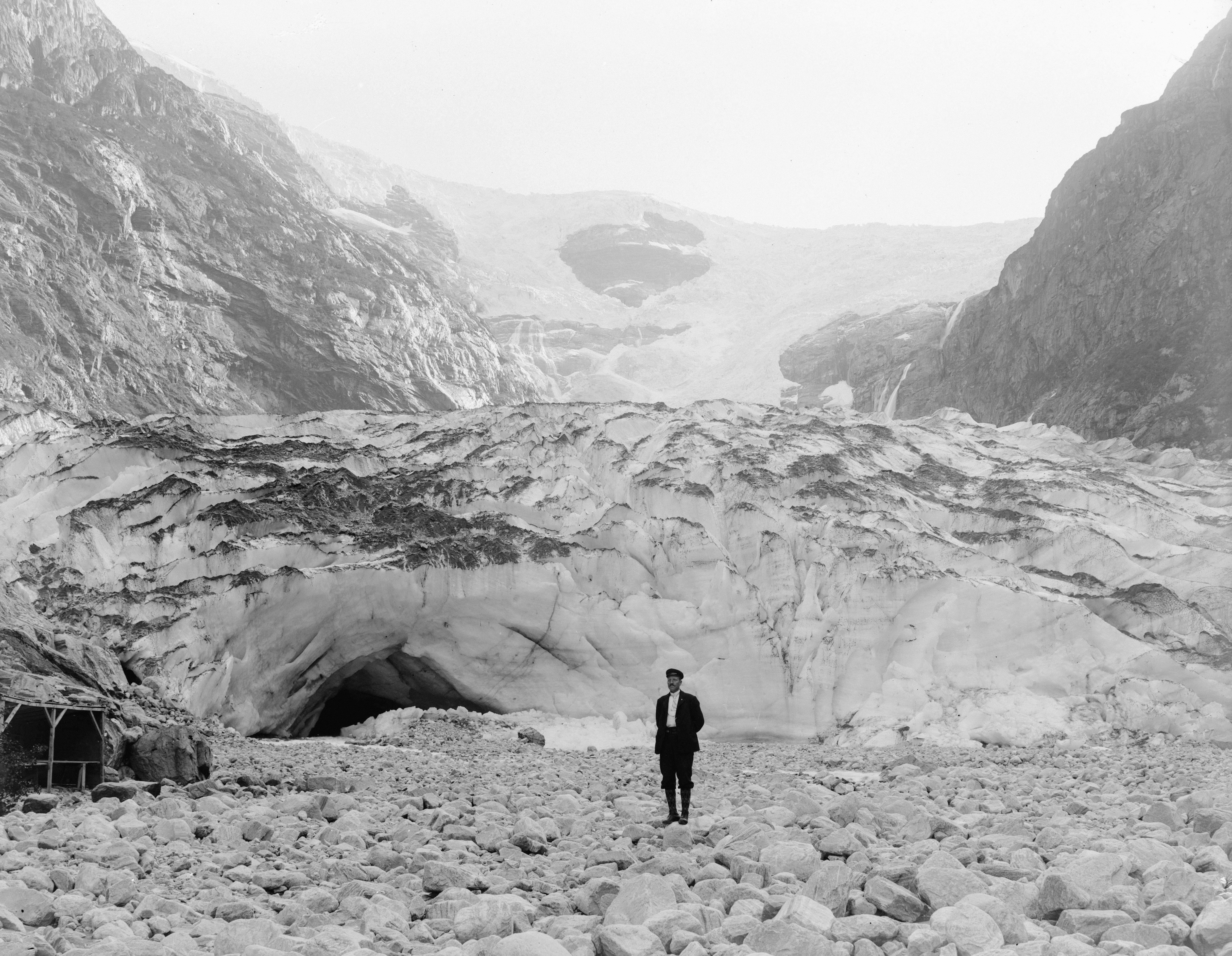 Black-and-white photograph of a lone man standing on a rocky glacier landscape with a dark cave opening in the background.