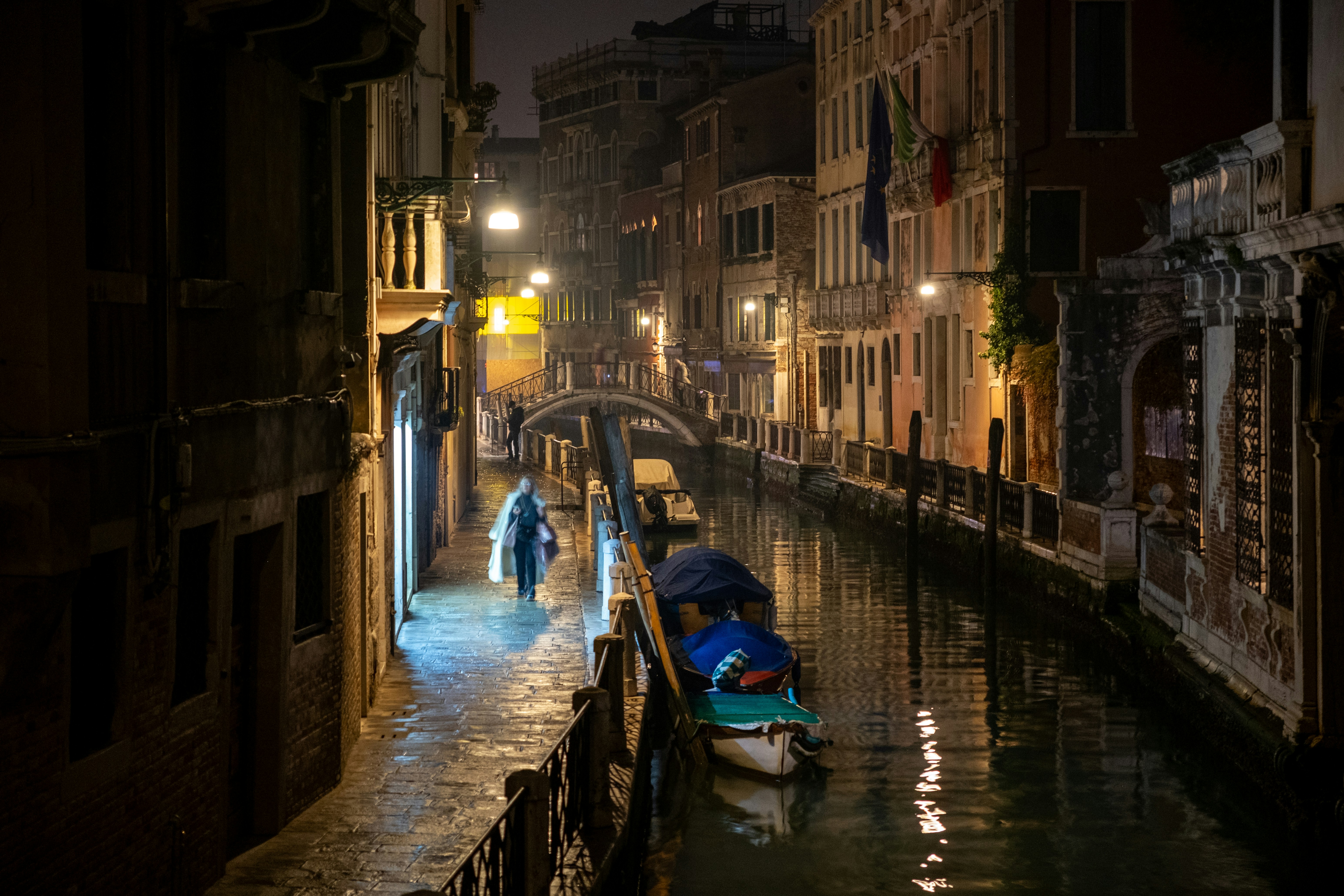 A serene canal scene in Venice at night, featuring dimly lit buildings and a solitary figure walking along the water's edge. The calm water reflects the ambient light, enhancing the tranquil atmosphere.