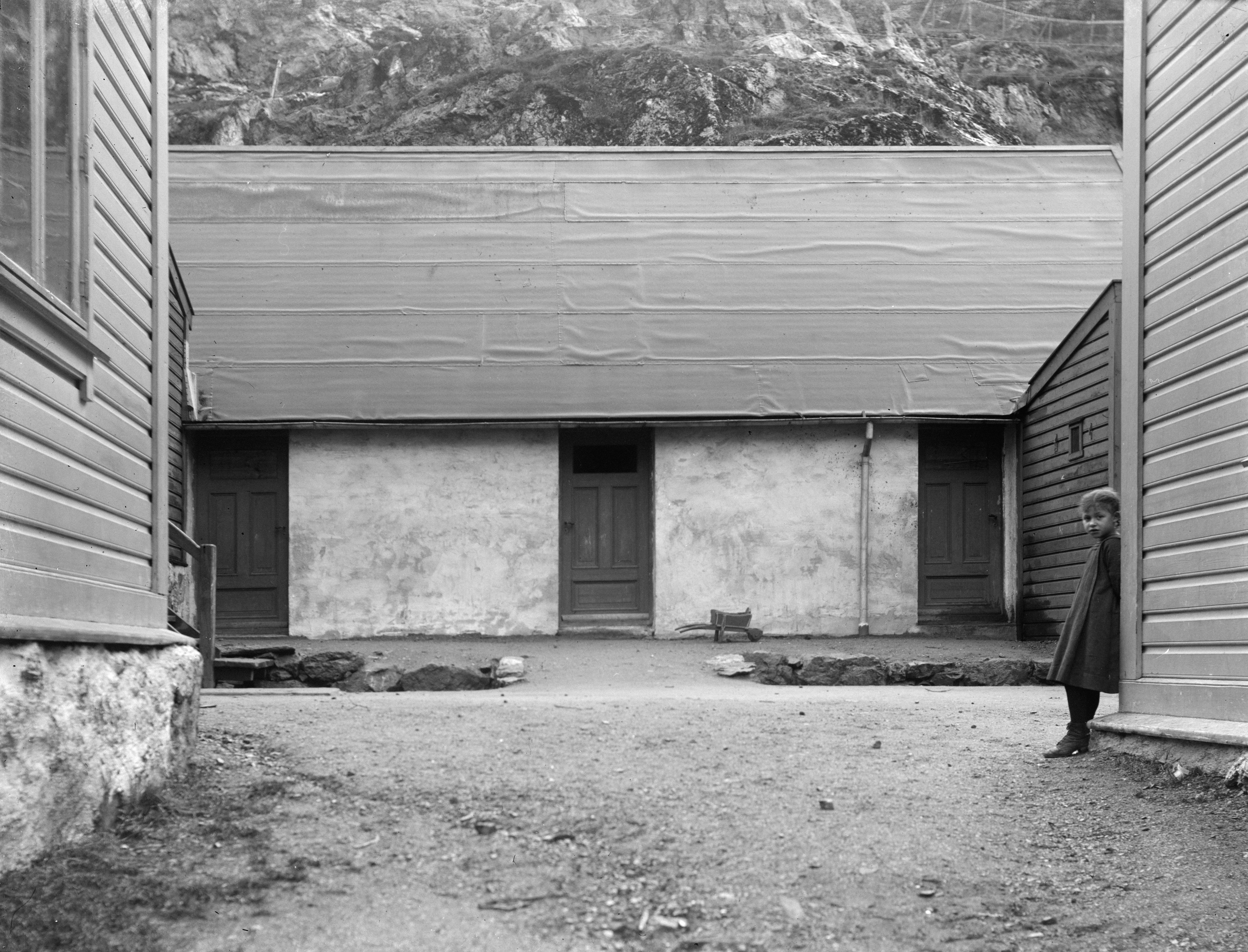 Monochrome photograph of a courtyard framed by two buildings, with a central doorway and a solitary figure on the right. The composition emphasizes austere architecture and restrained human presence.