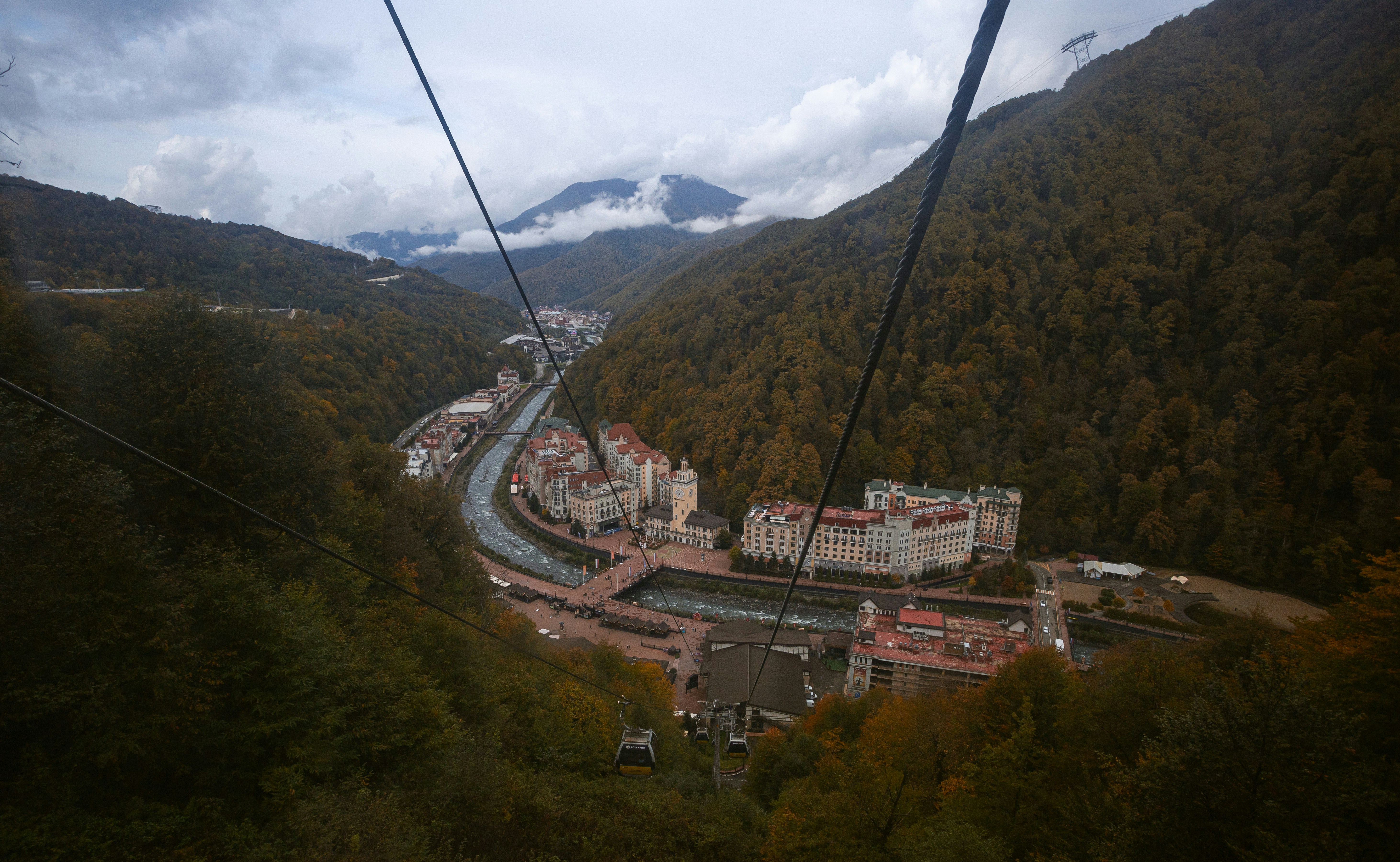 Aerial view of a town nestled in a valley with a winding river and autumn foliage, framed by forested mountains under an overcast sky.