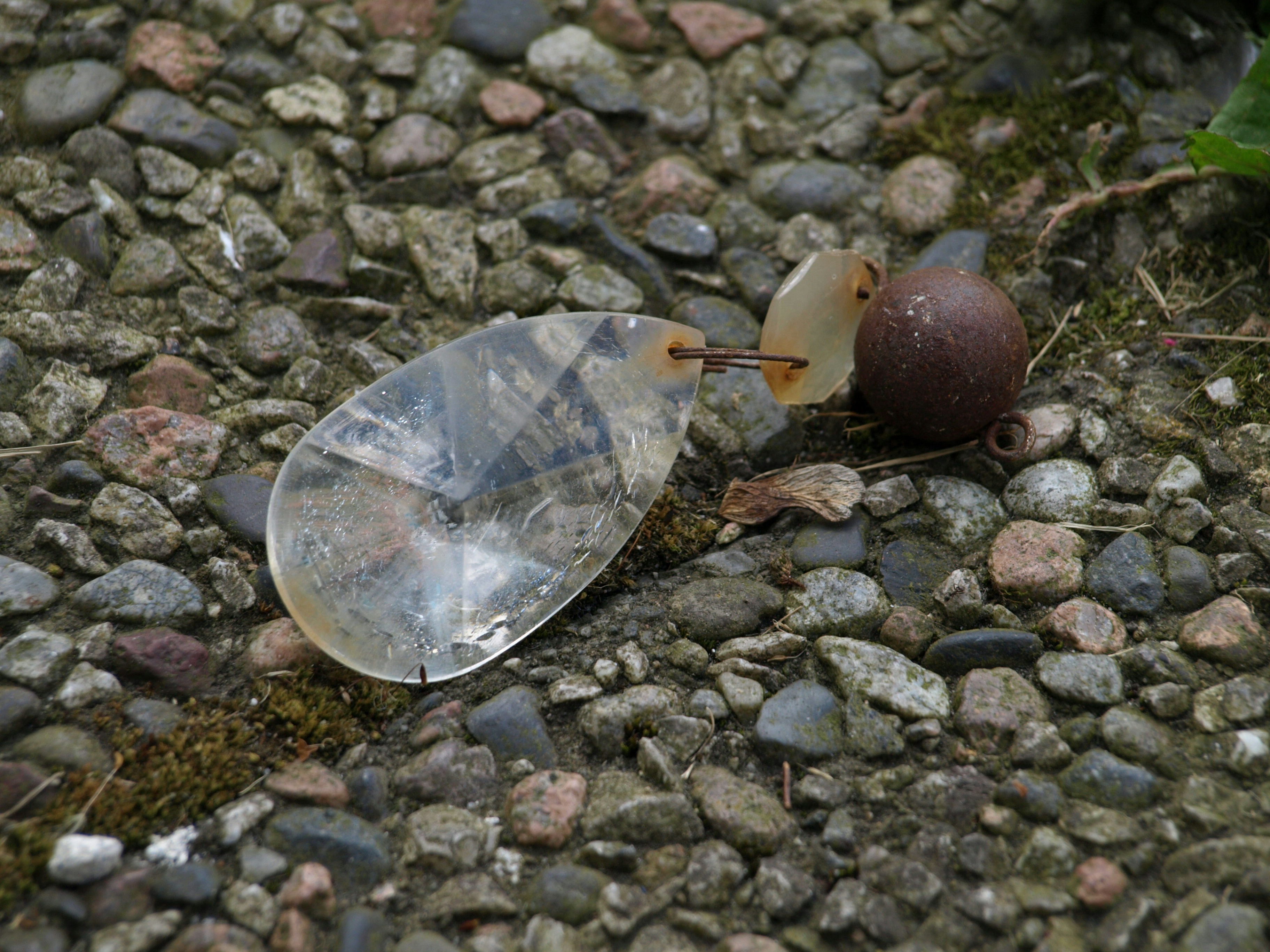Close-up photograph of a translucent, leaf-shaped shard resting on a gravel surface, with a small rusted bead nearby. The image emphasizes texture and contrast between the clear fragment and the rugged pebbles.