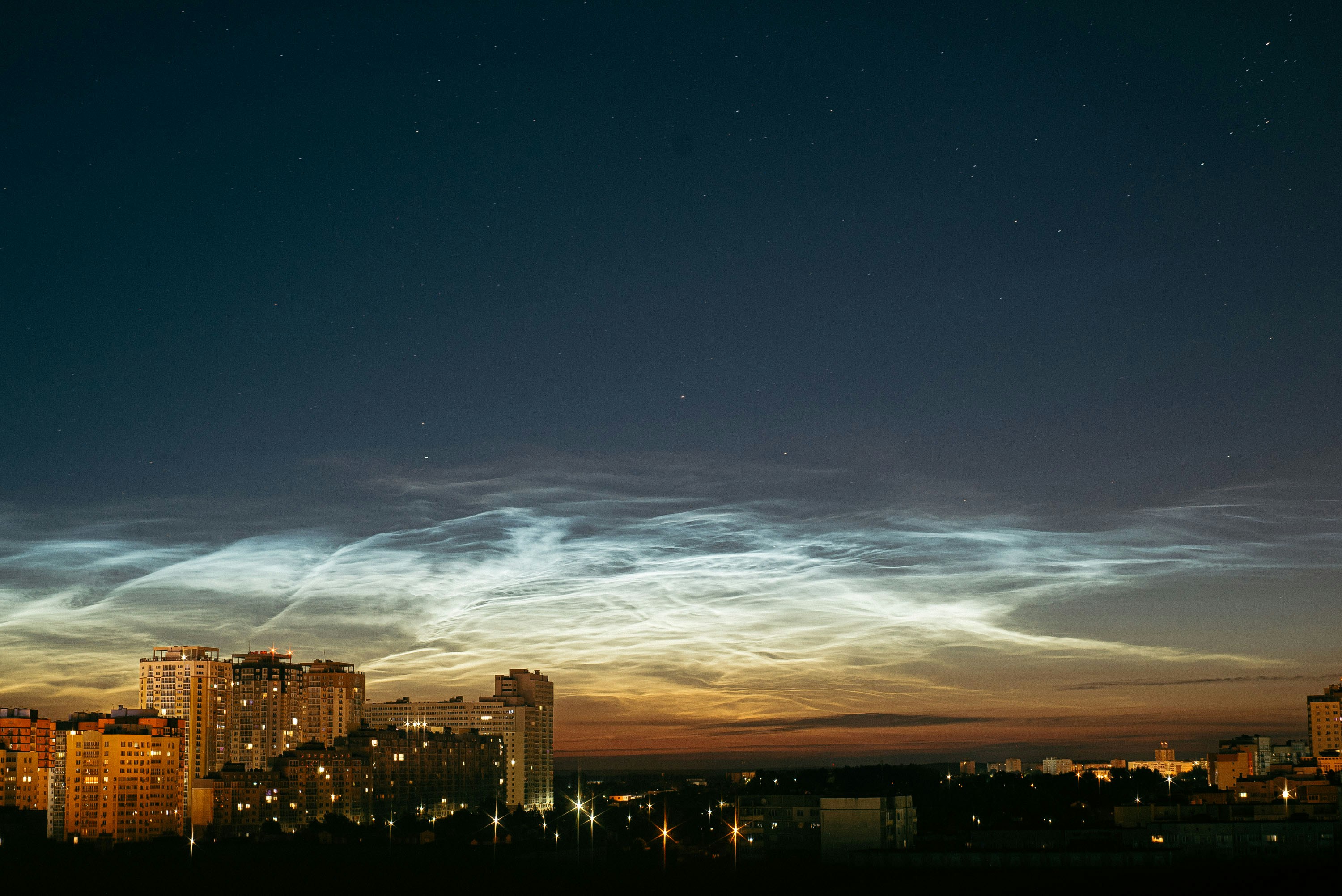 Cityscape at dusk with faint noctilucent clouds against a darkening sky.