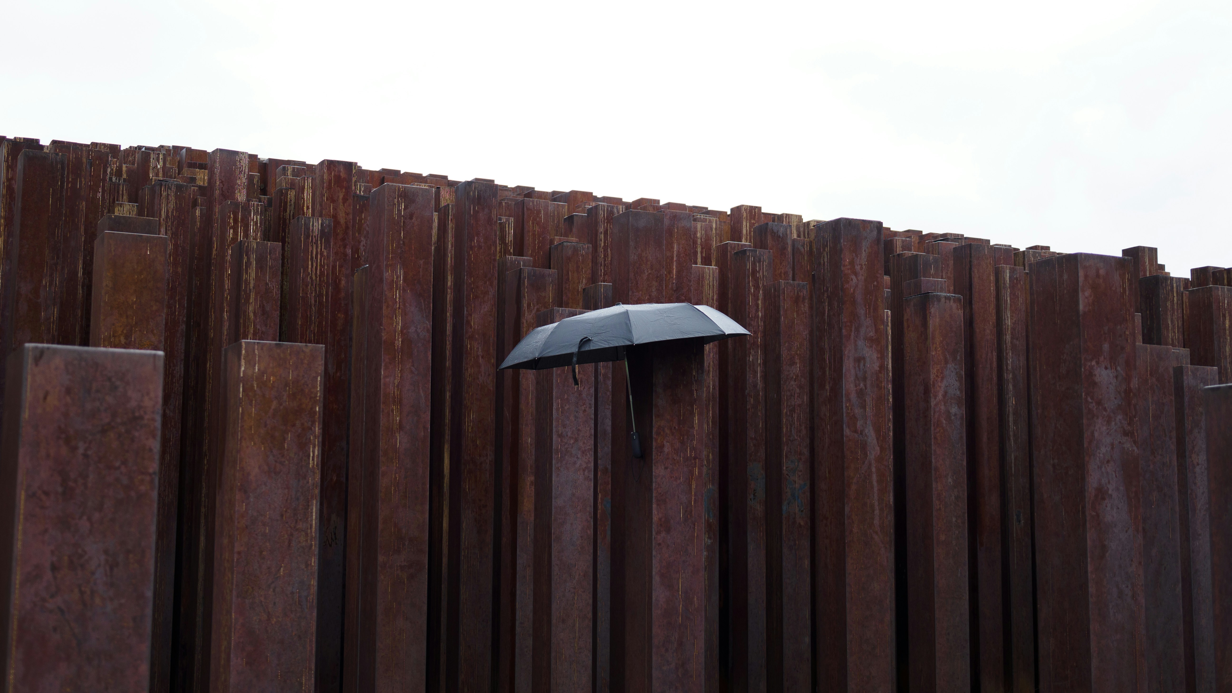 A black umbrella stands among vertical, rusted steel columns in a stark, industrial landscape.