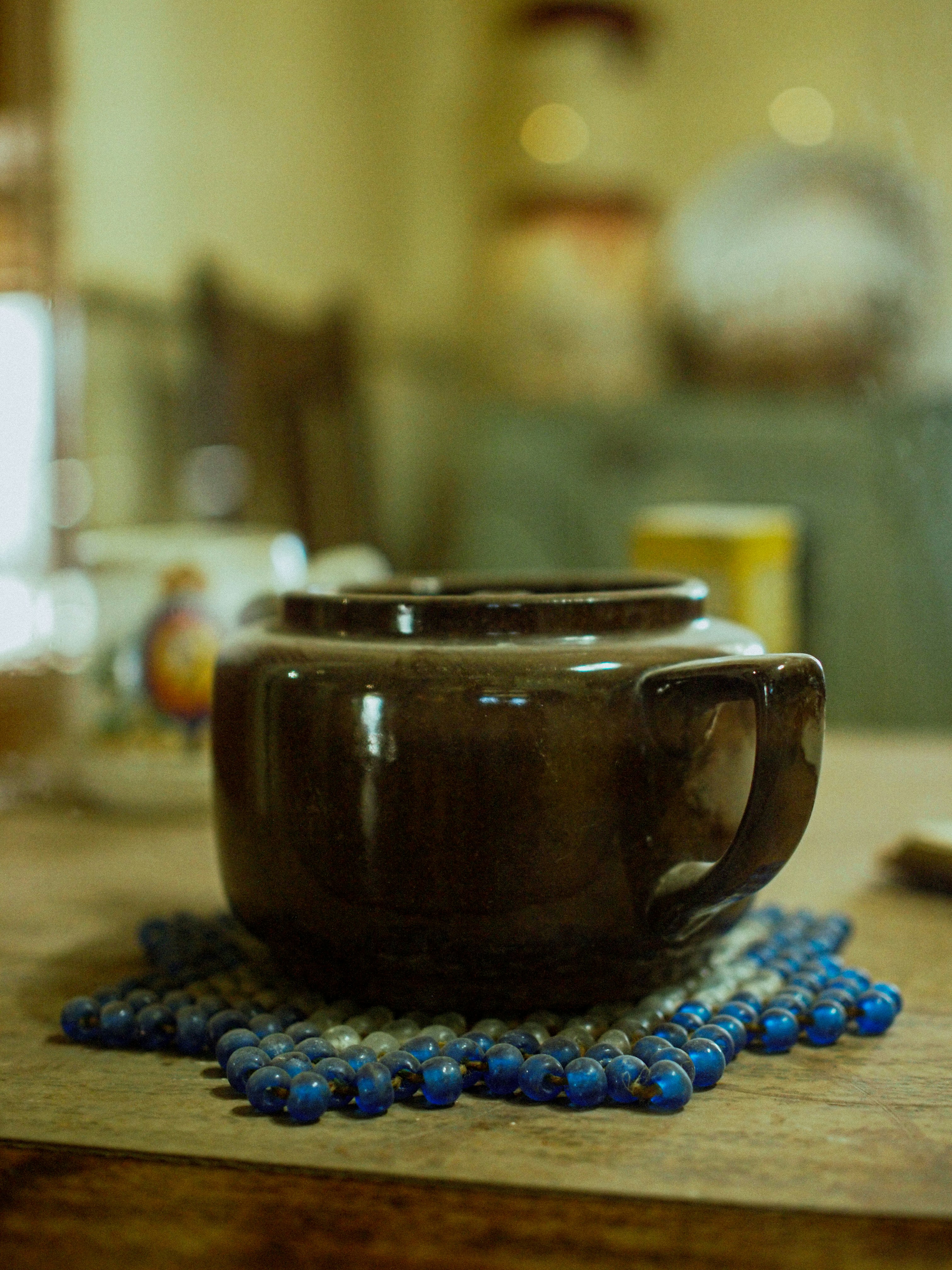 A dark ceramic teapot rests on a vibrant blue beaded mat, surrounded by a warm, softly blurred interior setting.