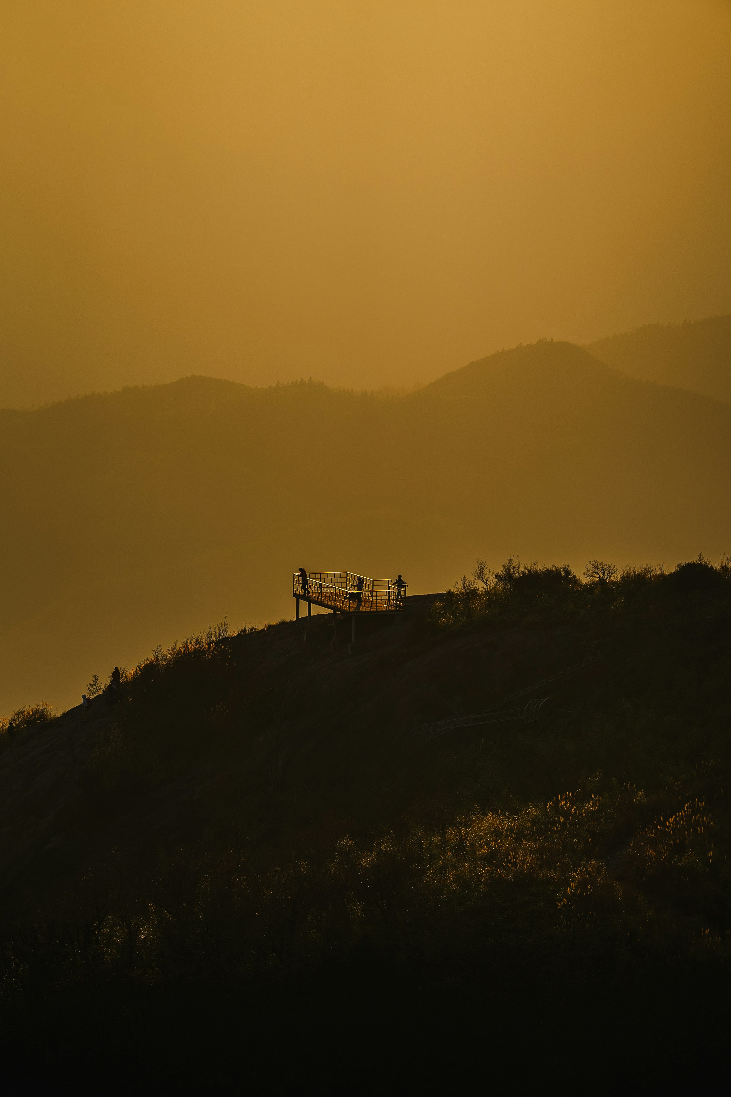 A wooden observation deck stands alone on a mountain ridge, surrounded by softly layered hills bathed in golden light during sunset.