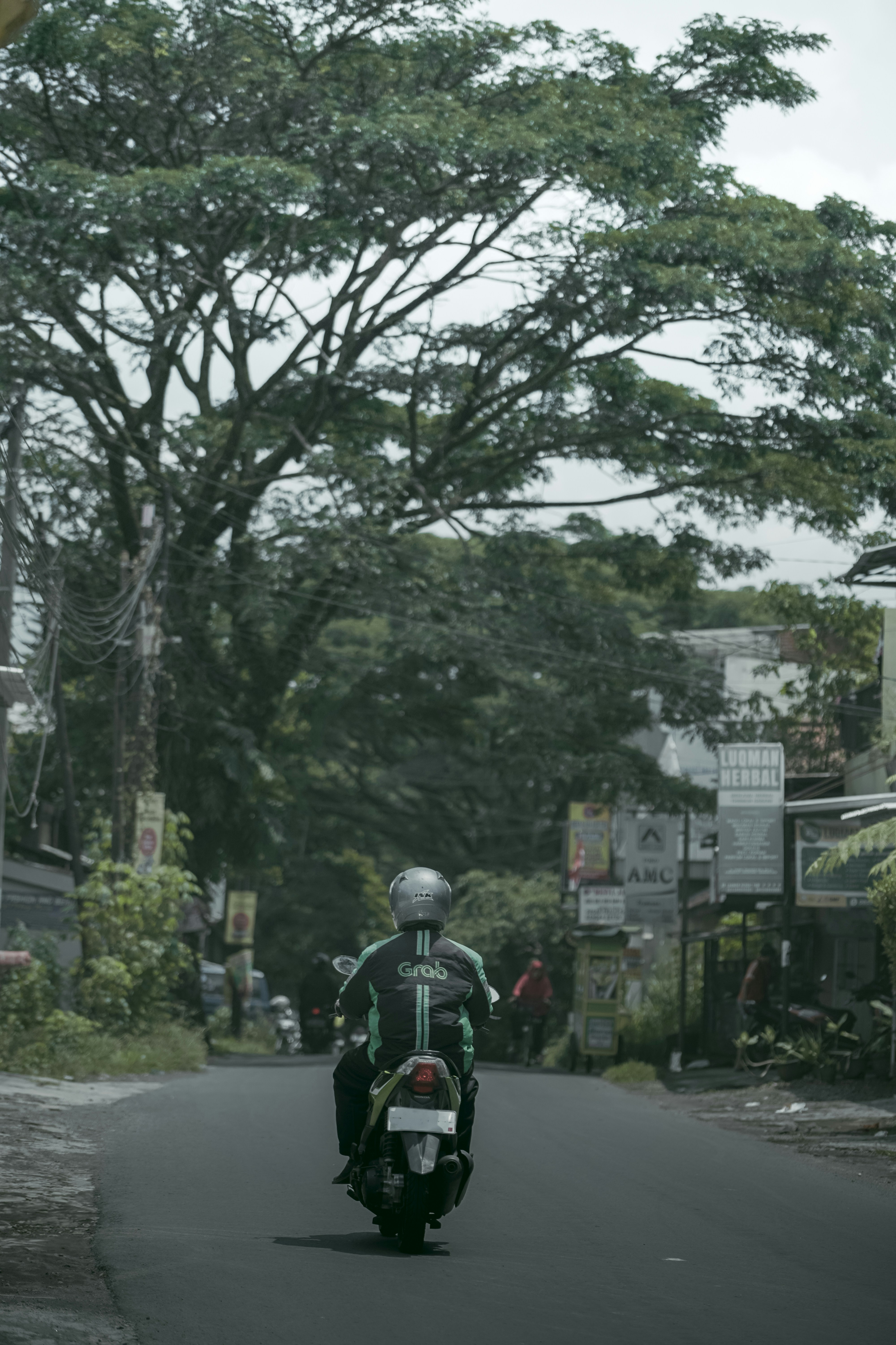 A motorbike rider in a green jacket navigates a quiet street lined with lush trees and local shops. The scene captures the essence of everyday life in a vibrant community.
