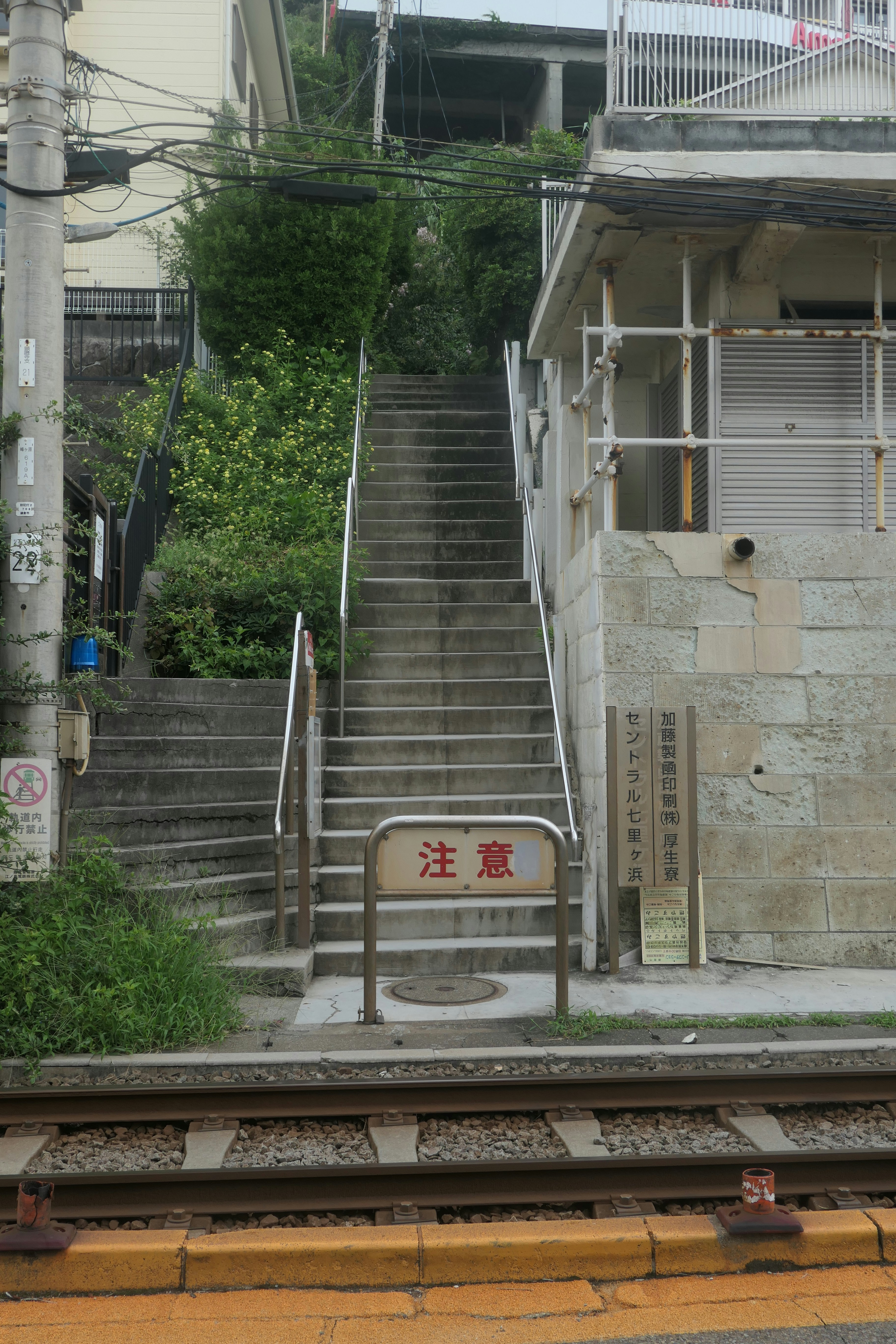 Concrete stairway rises between a weathered building and lush greenery, flanking the railway tracks in the foreground. A caution sign and scaffolding hint at maintenance in a restrained urban setting.