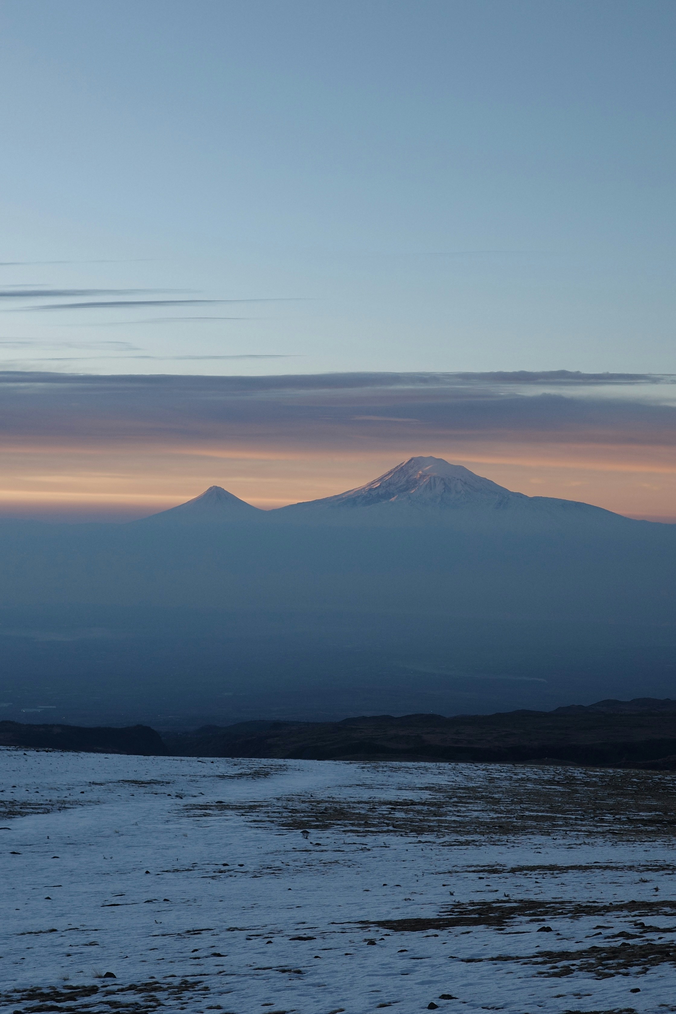 Two majestic mountains shrouded in mist during twilight, with a soft gradient of colors in the sky. Snow blankets the foreground, enhancing the serene atmosphere.