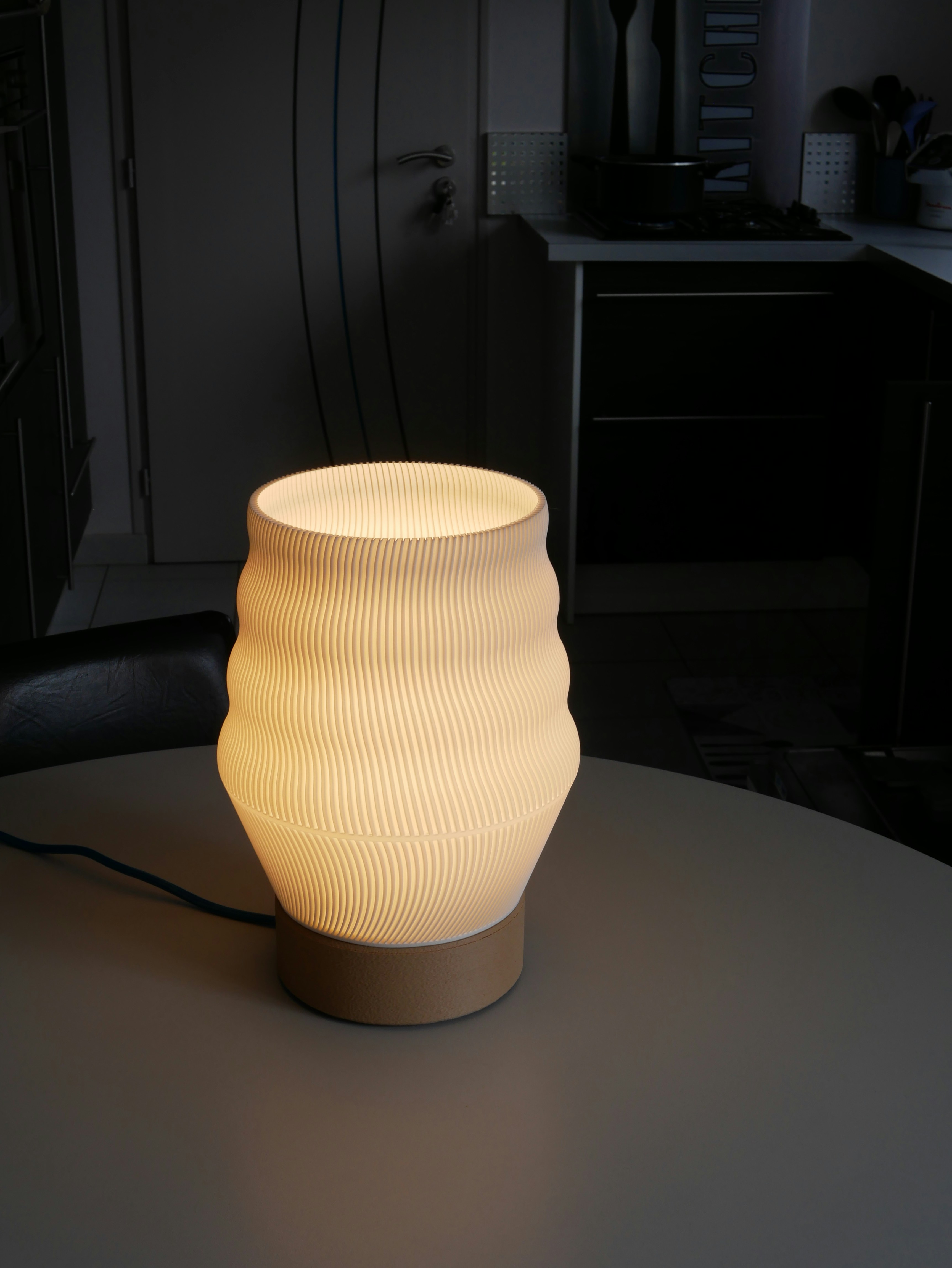 A 3D-printed lamp by HomaKraft, EProjets Lab, glowing warmly in a dark environment. The lamp, with its long minimalistic design, stands on a white round table, casting a soft light in the room. A black leather chair back and a white door in the background create a clean contrast with the lamp's warm glow. The shot is taken from a half-front, half-overhead angle, showcasing the lamp's intricate details and the cozy, inviting ambiance.
