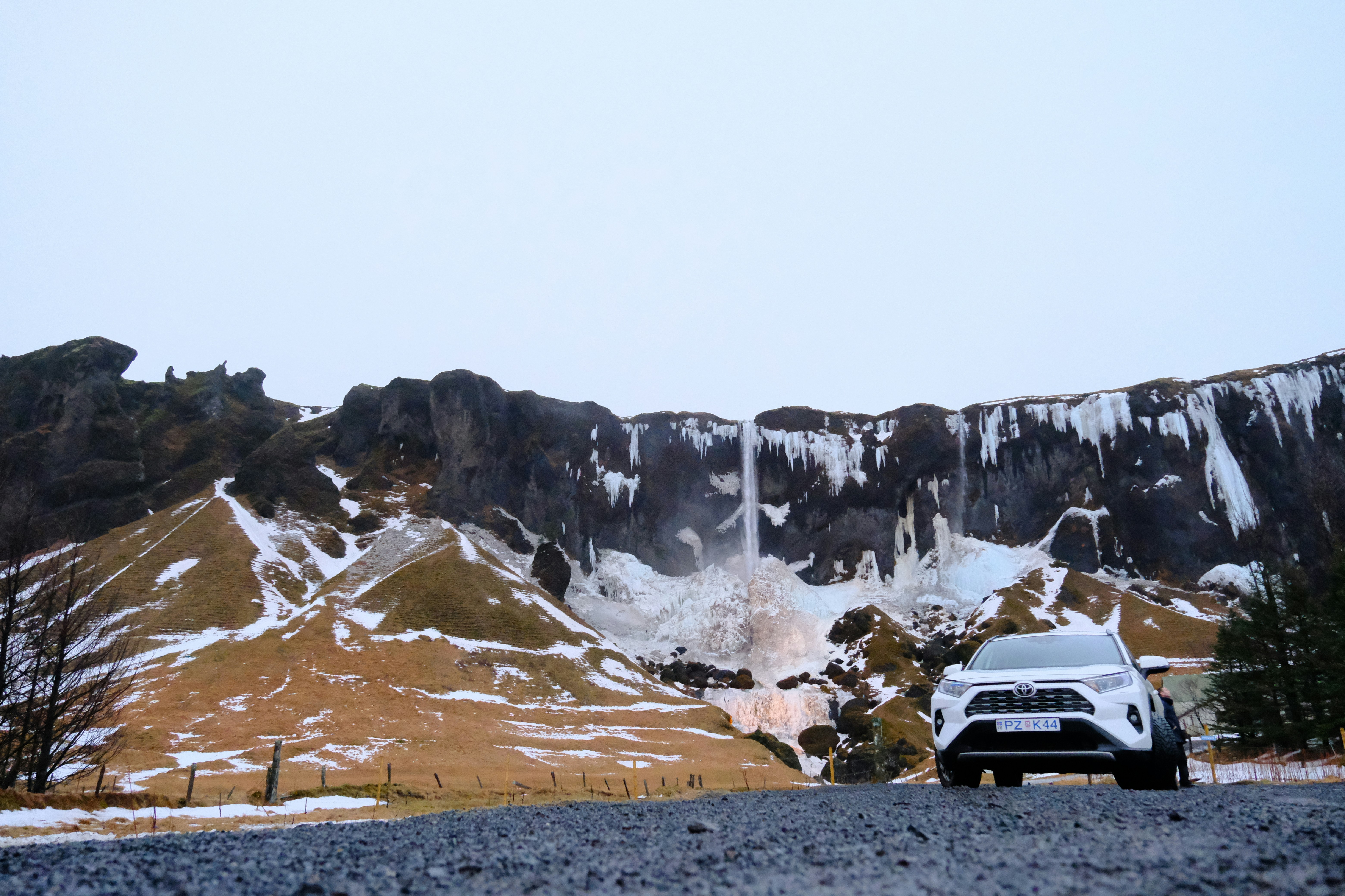 White truck on a snow-dusted road beneath rugged cliffs and cascading waterfall.