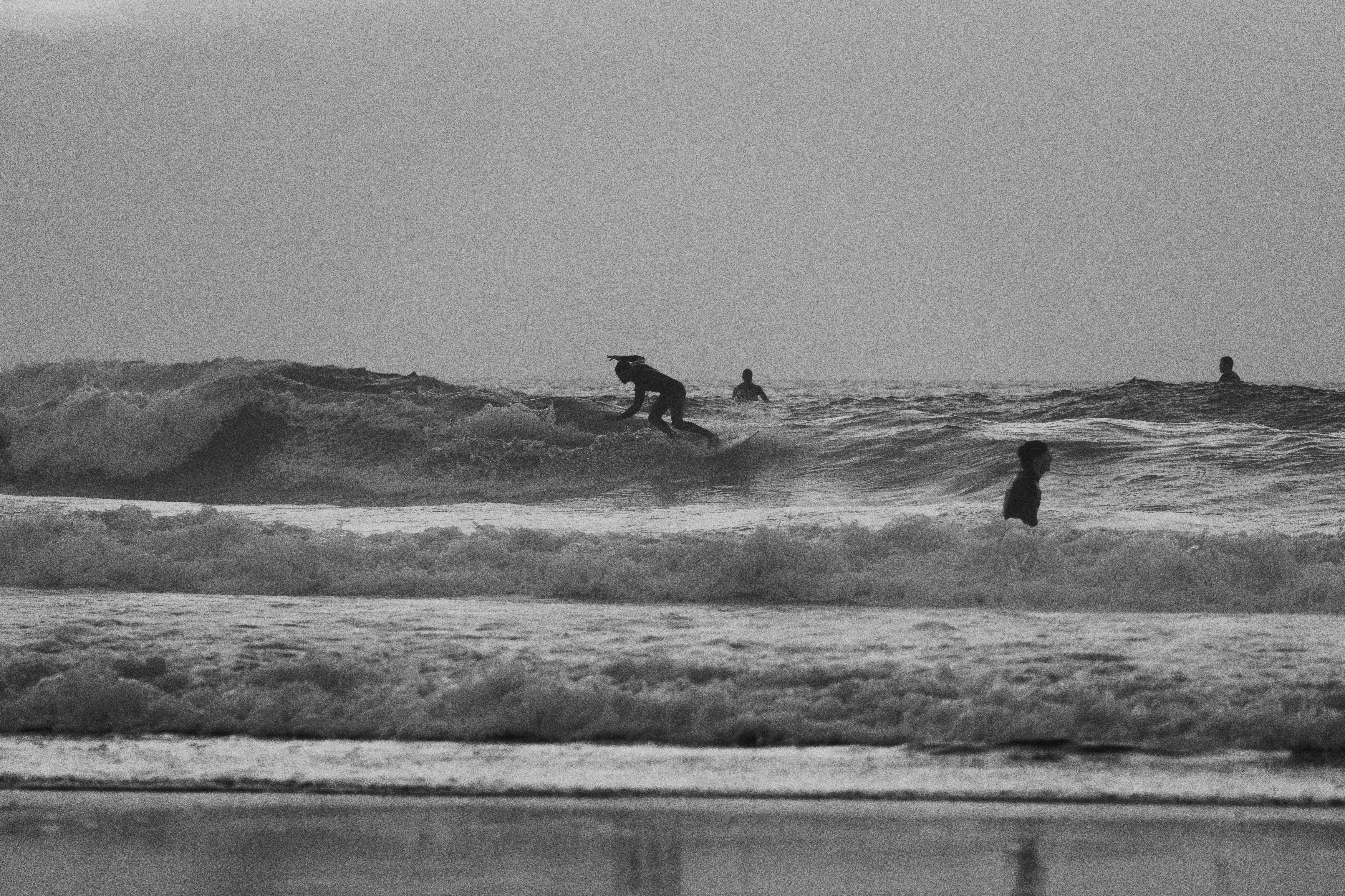 Surfers navigate rolling waves under a dim sky, captured in monochrome.