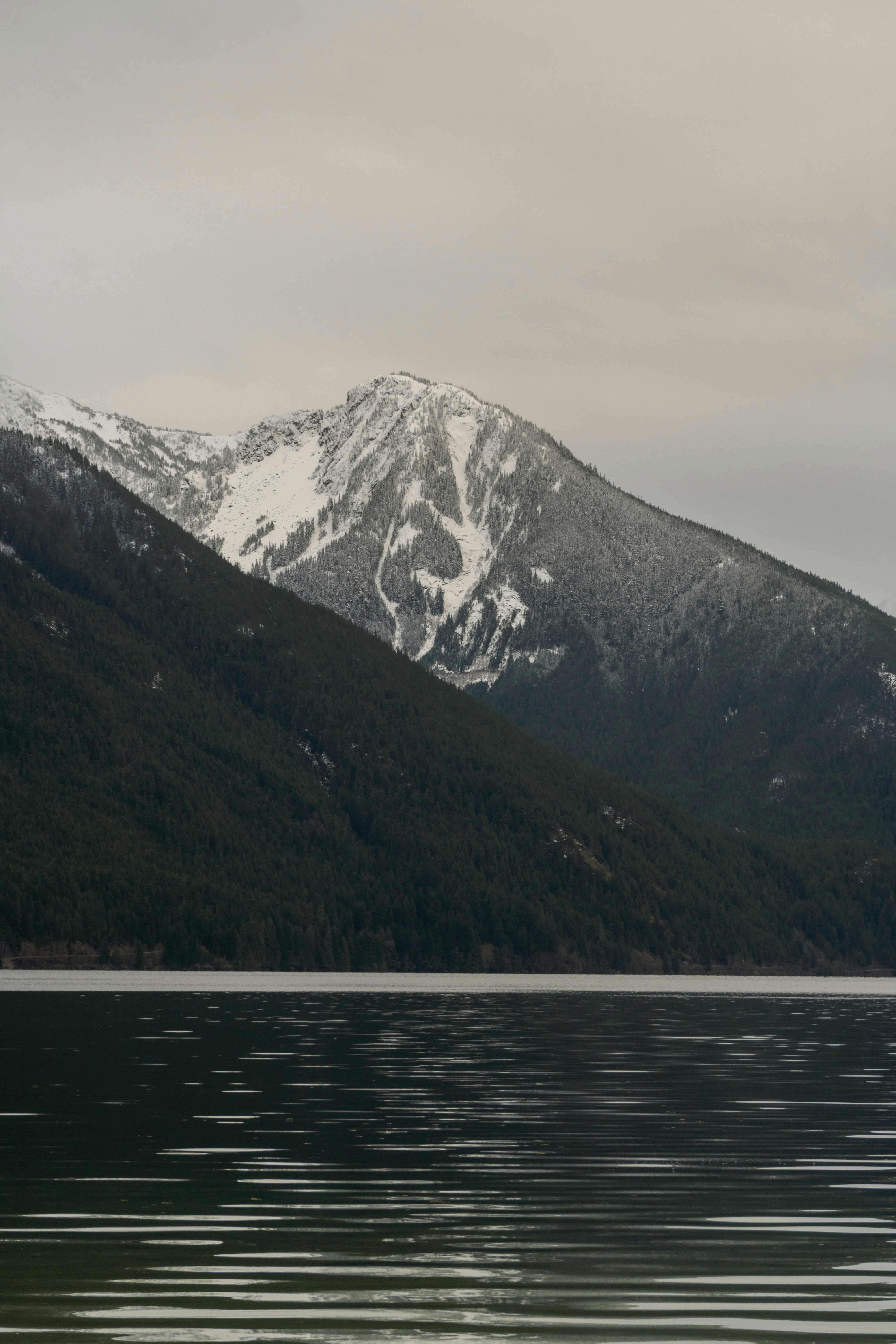 Snow-capped mountains rise above a tranquil lake, reflecting the muted tones of a cloudy sky. The serene landscape captures the essence of winter's embrace.