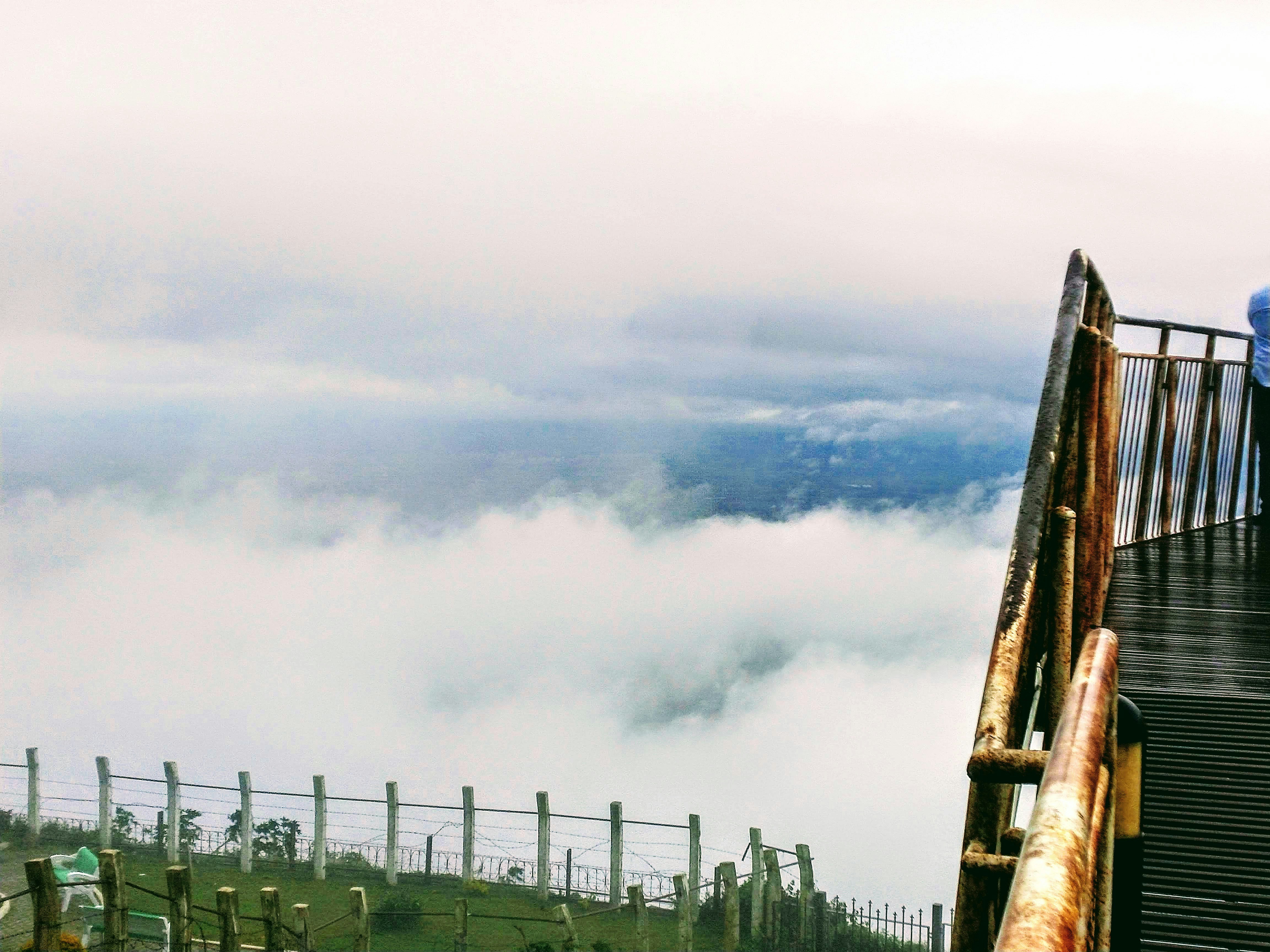 Mist shrouds a mountainous edge as a weathered wooden boardwalk extends toward the horizon, with railing fading into the cloudscape.