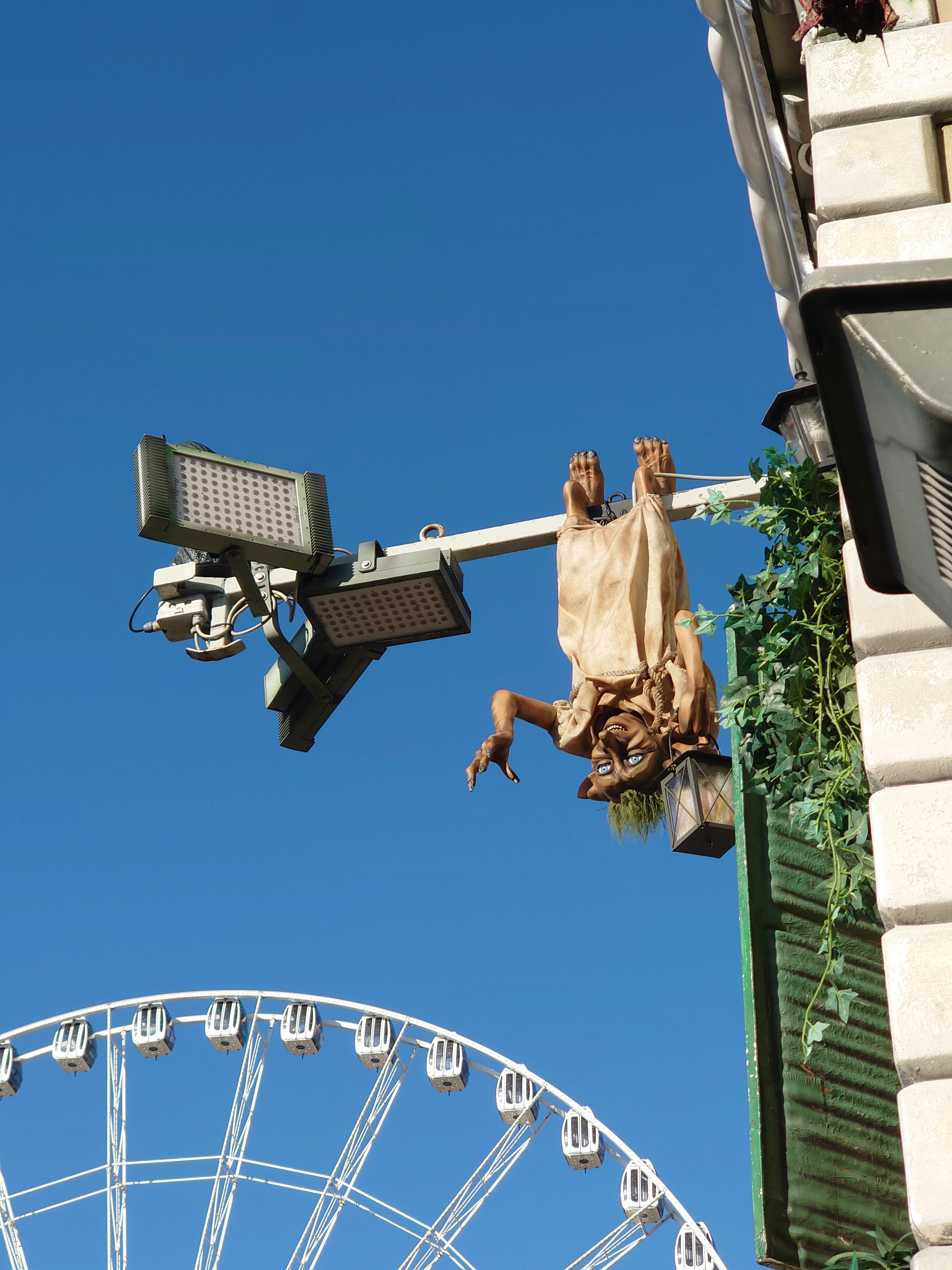 Upside-down sculpture suspended from a street beam against a vivid blue sky; a partial ferris wheel arc and building facade frame the scene.