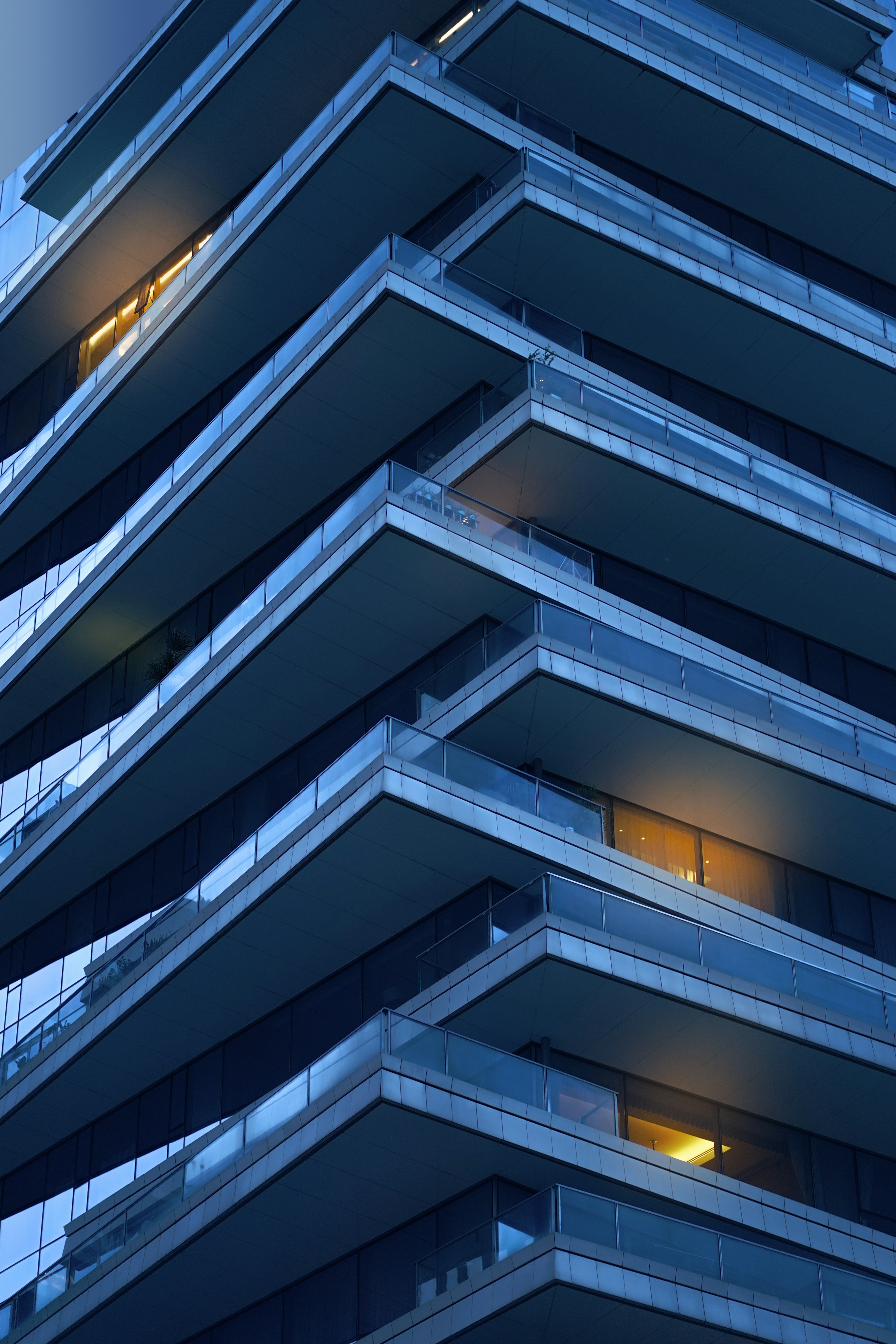 Modern residential building with geometric balconies at twilight, featuring cool blue tones and warm window lights.