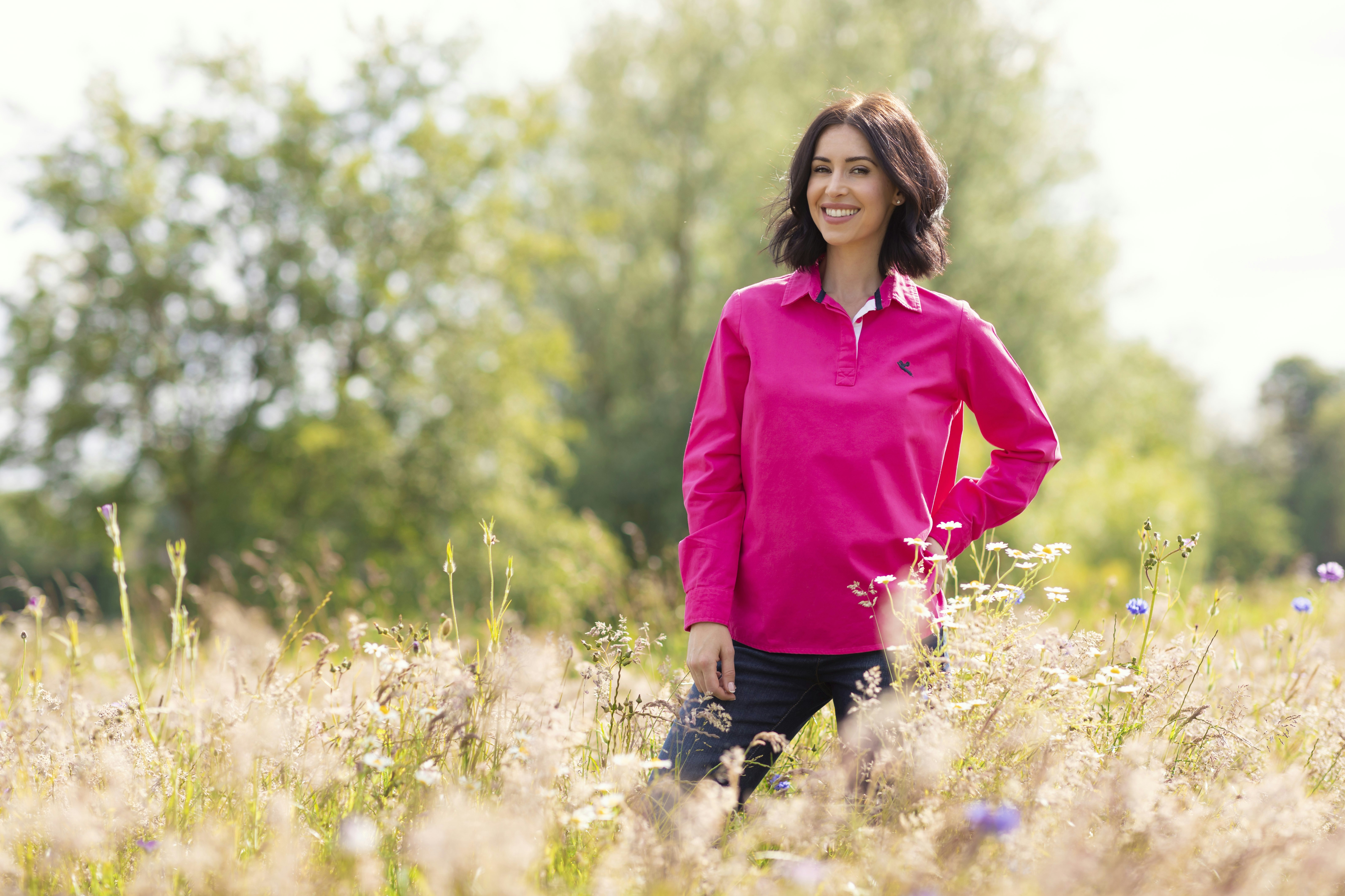 Lady wearing a bright pink long sleeve top with a collar standing amongst long grasses in the British countryside. | A woman standing in a field of tall grass