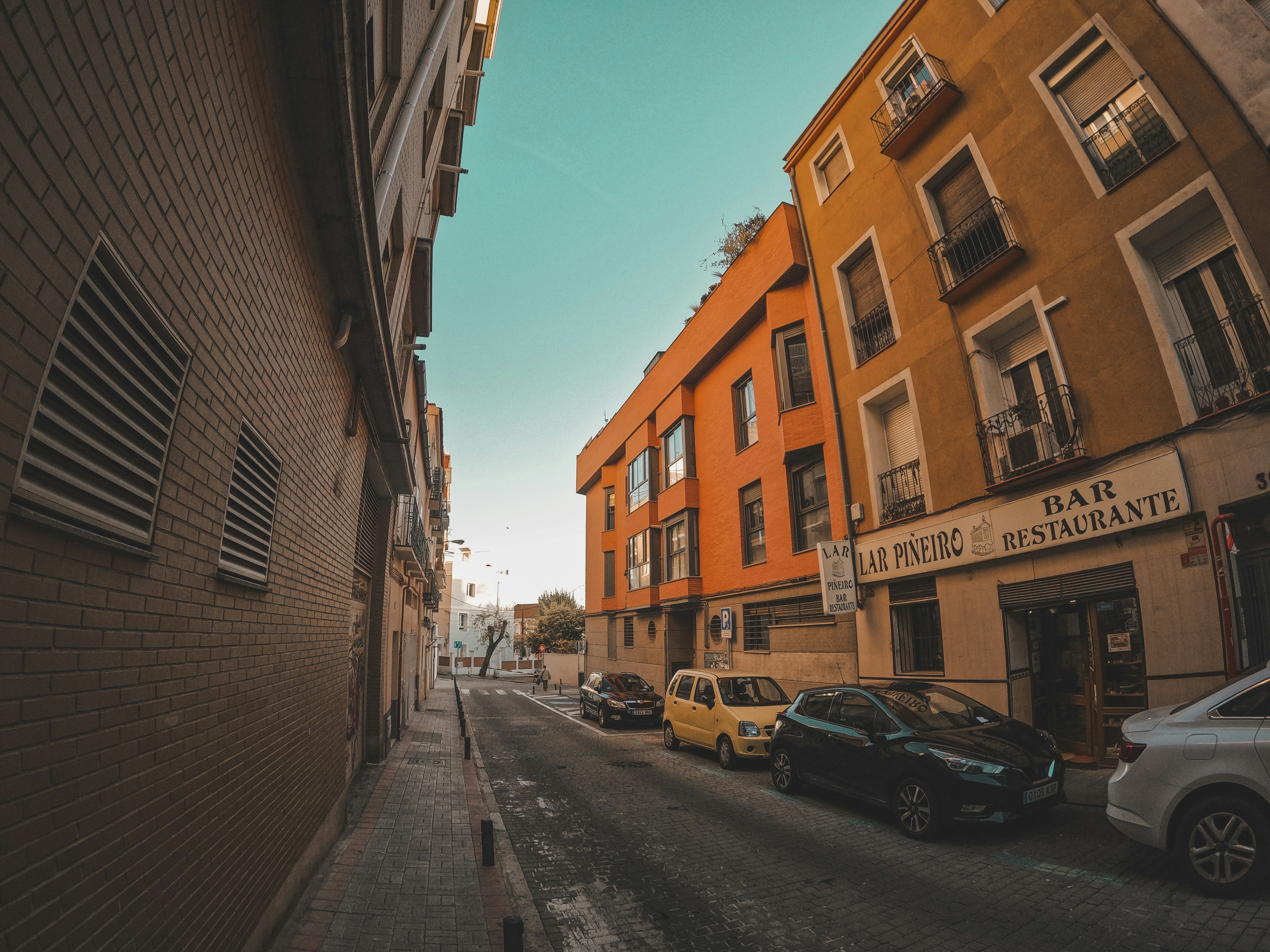 Quiet urban street with vibrant orange and yellow buildings under a soft cyan sky at golden hour.