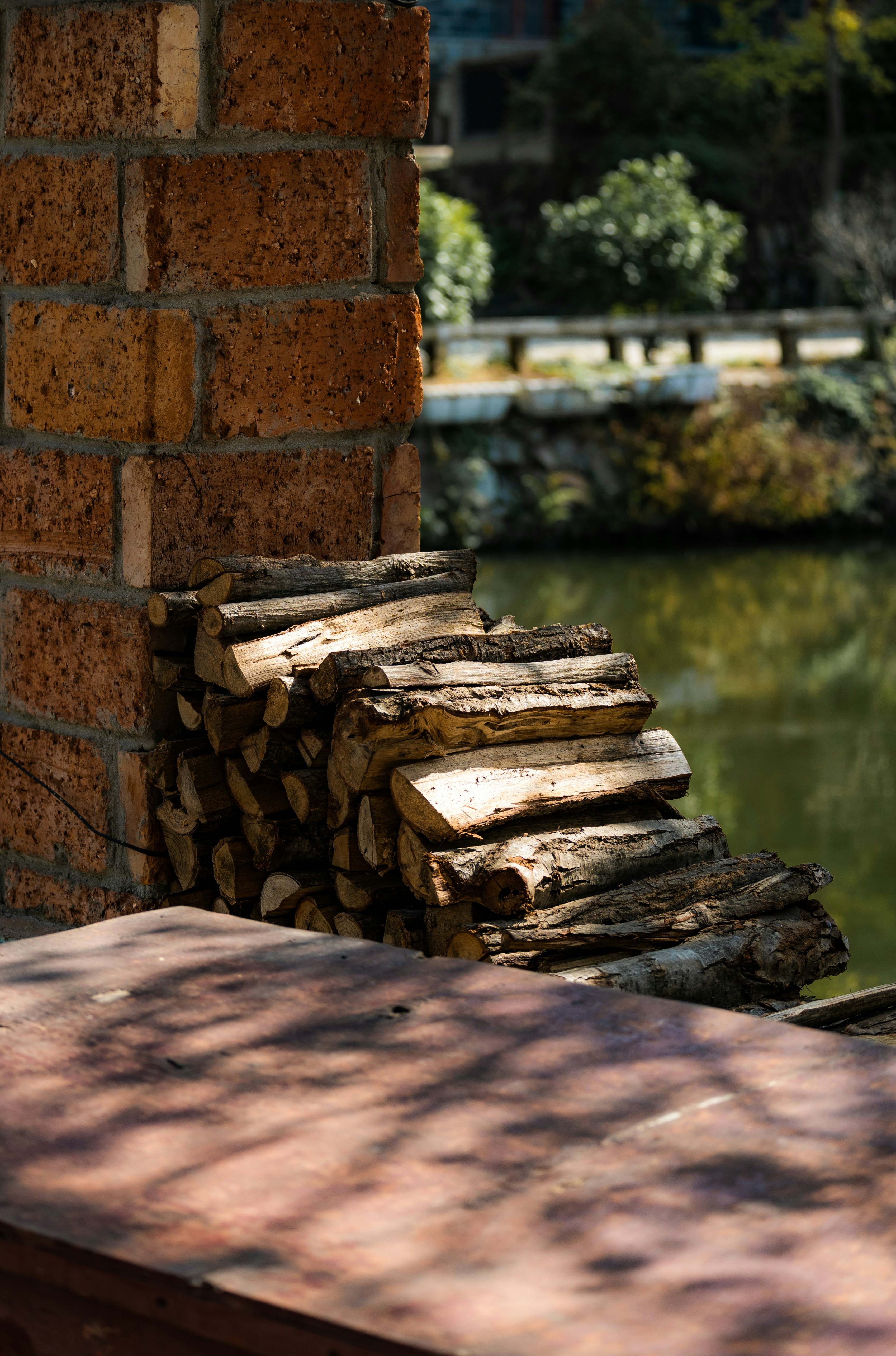 Quiet Tension: Stacked Firewood at a Brick Pillar by a Reflective Pond