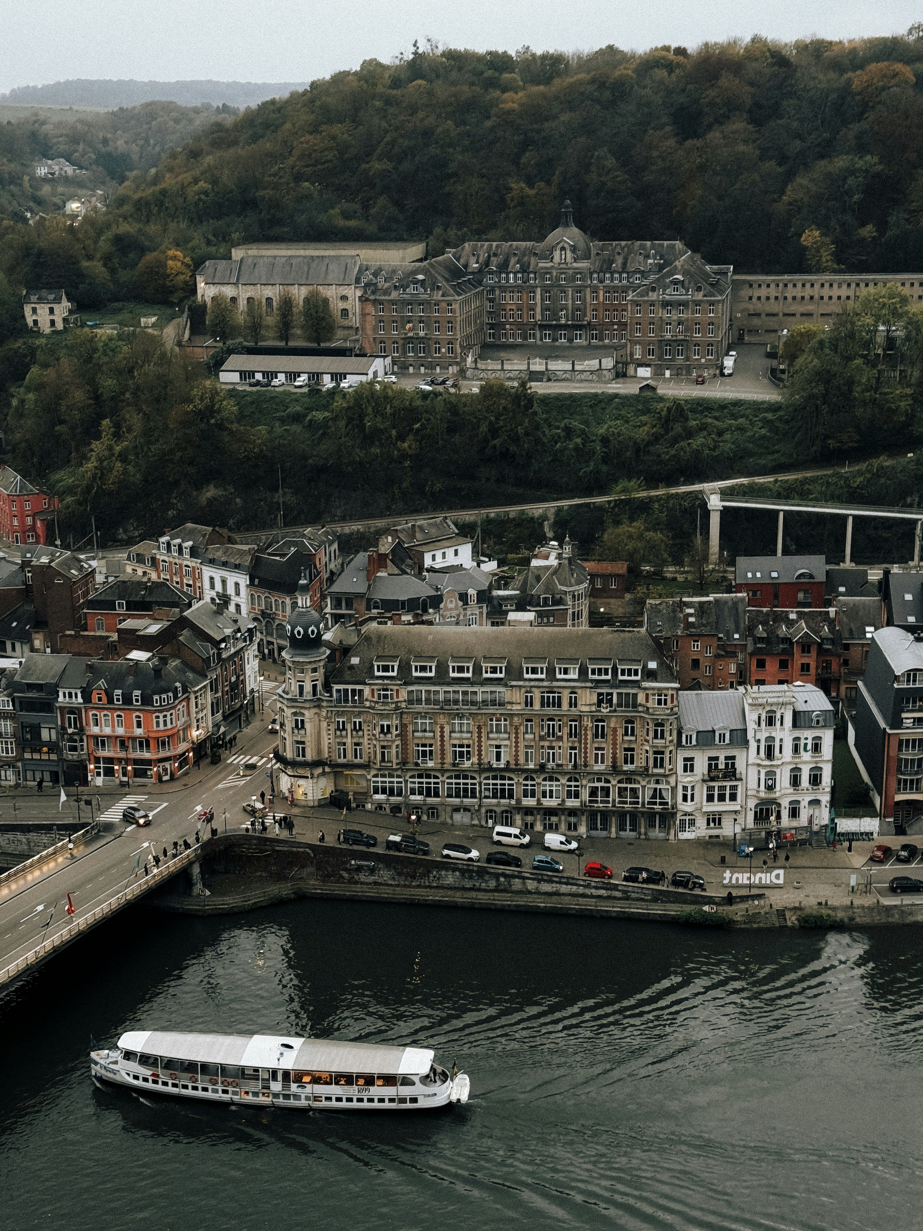 A boat glides along a river lined with historic buildings and lush, green hills under an overcast sky.