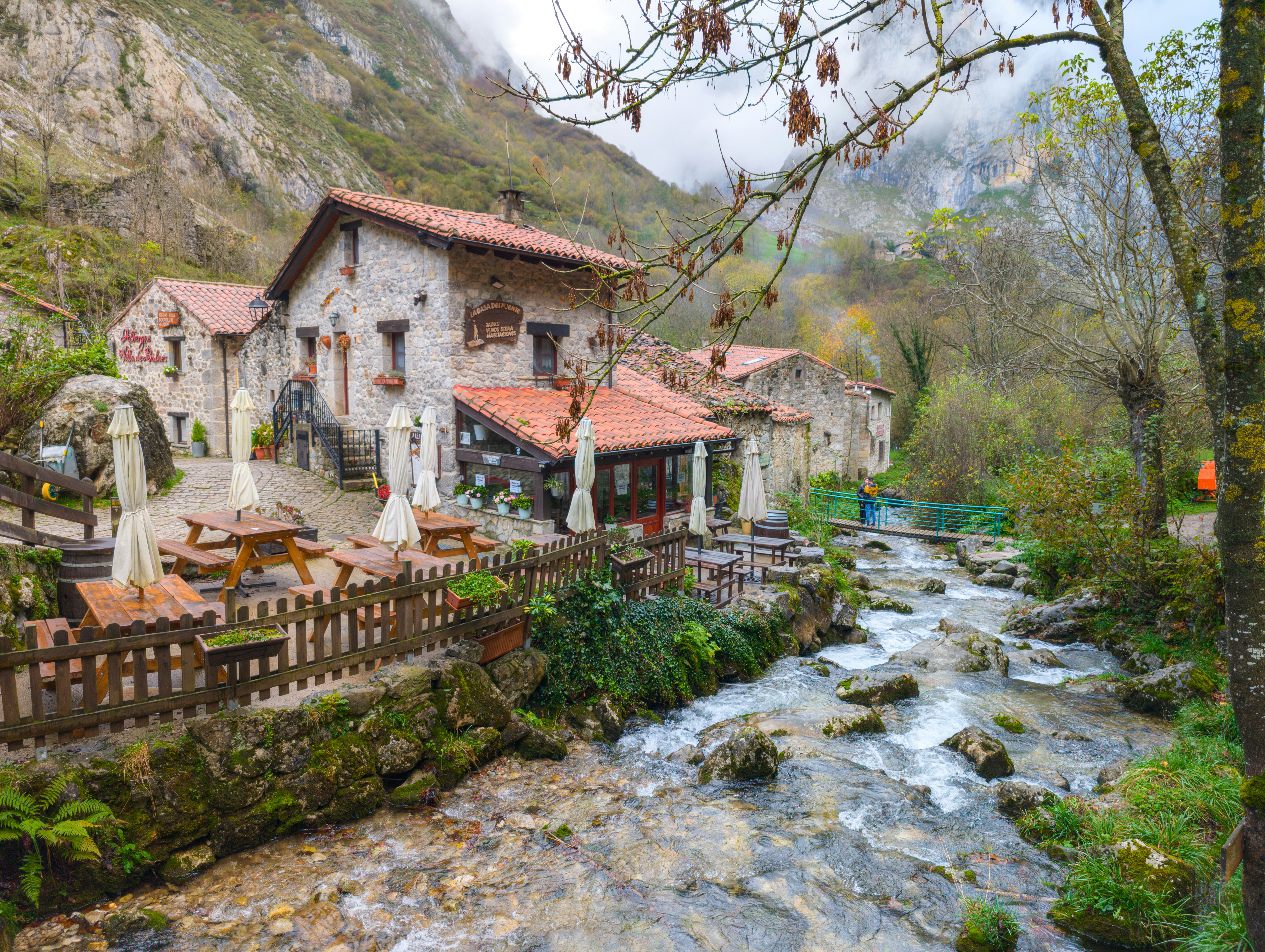 Stone cottage with red roofs nestled beside a flowing stream in a mountainous landscape.