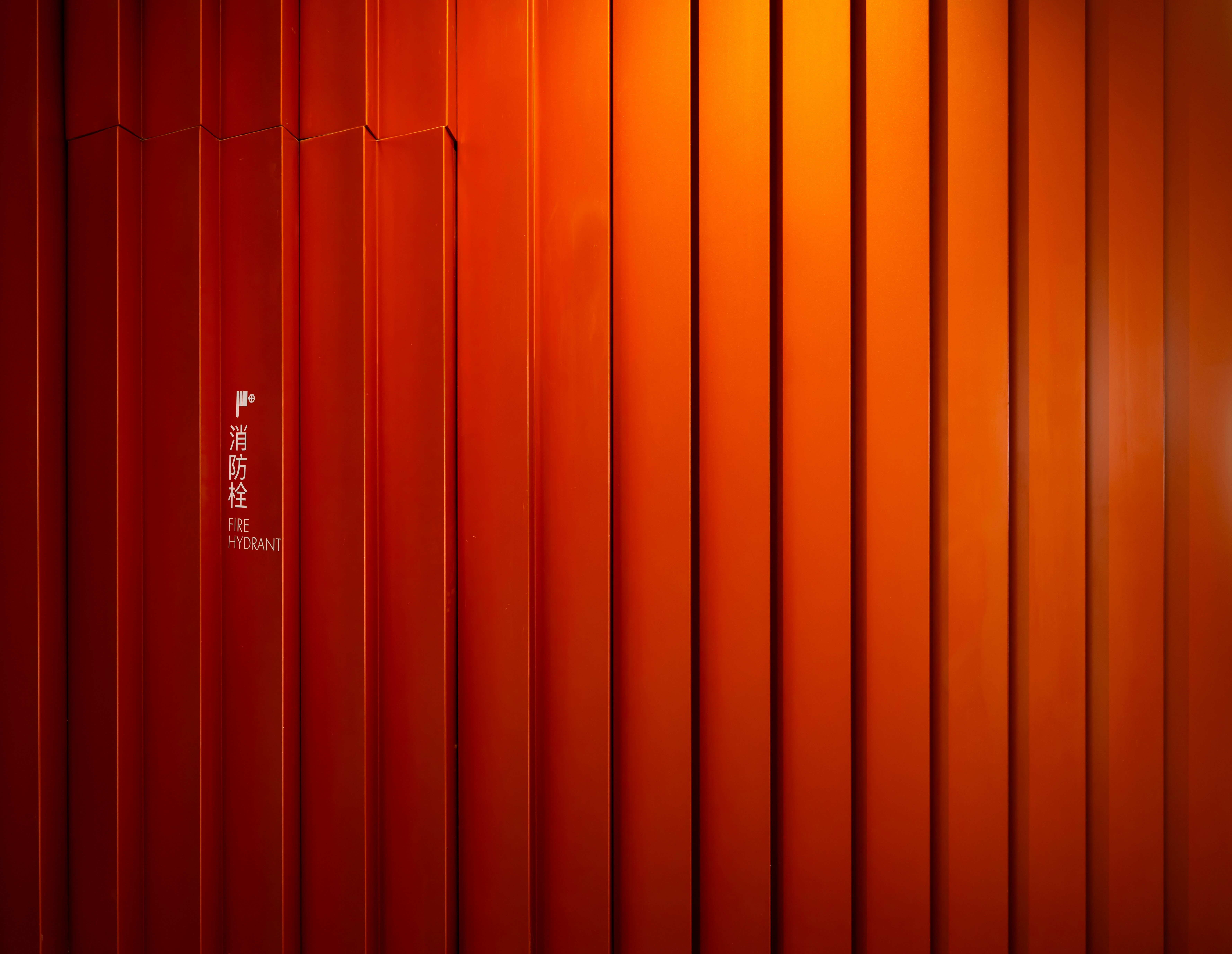 Close-up of vertical orange panels creating a geometric wall, with a small white fire hydrant sign along the left edge.