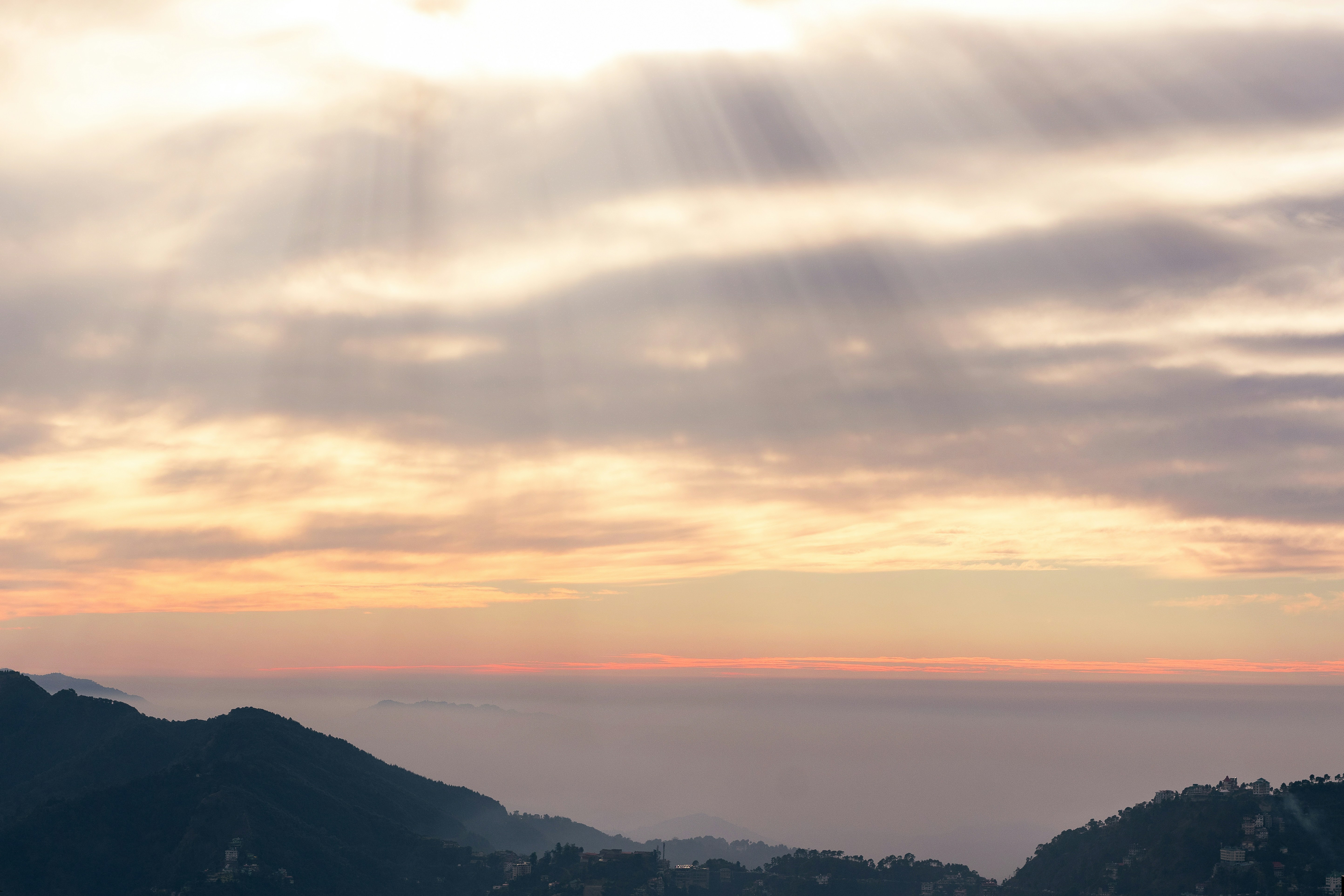 Sunlight pierces through layered clouds above a mountain range at sunset.