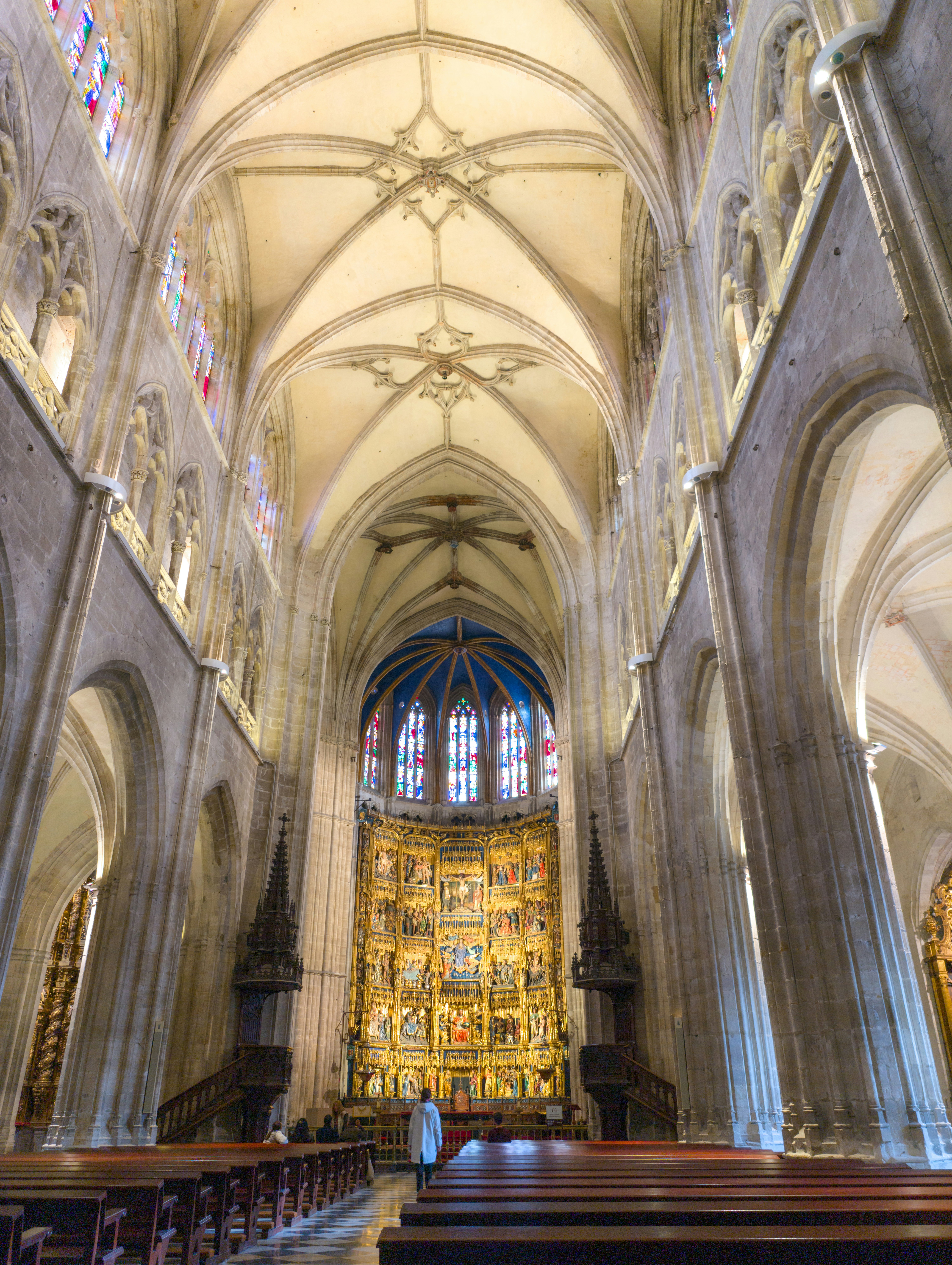 Intricate stone arches and vibrant stained glass illuminate the vast interior of a Gothic cathedral, showcasing an ornate altarpiece at the focal point.