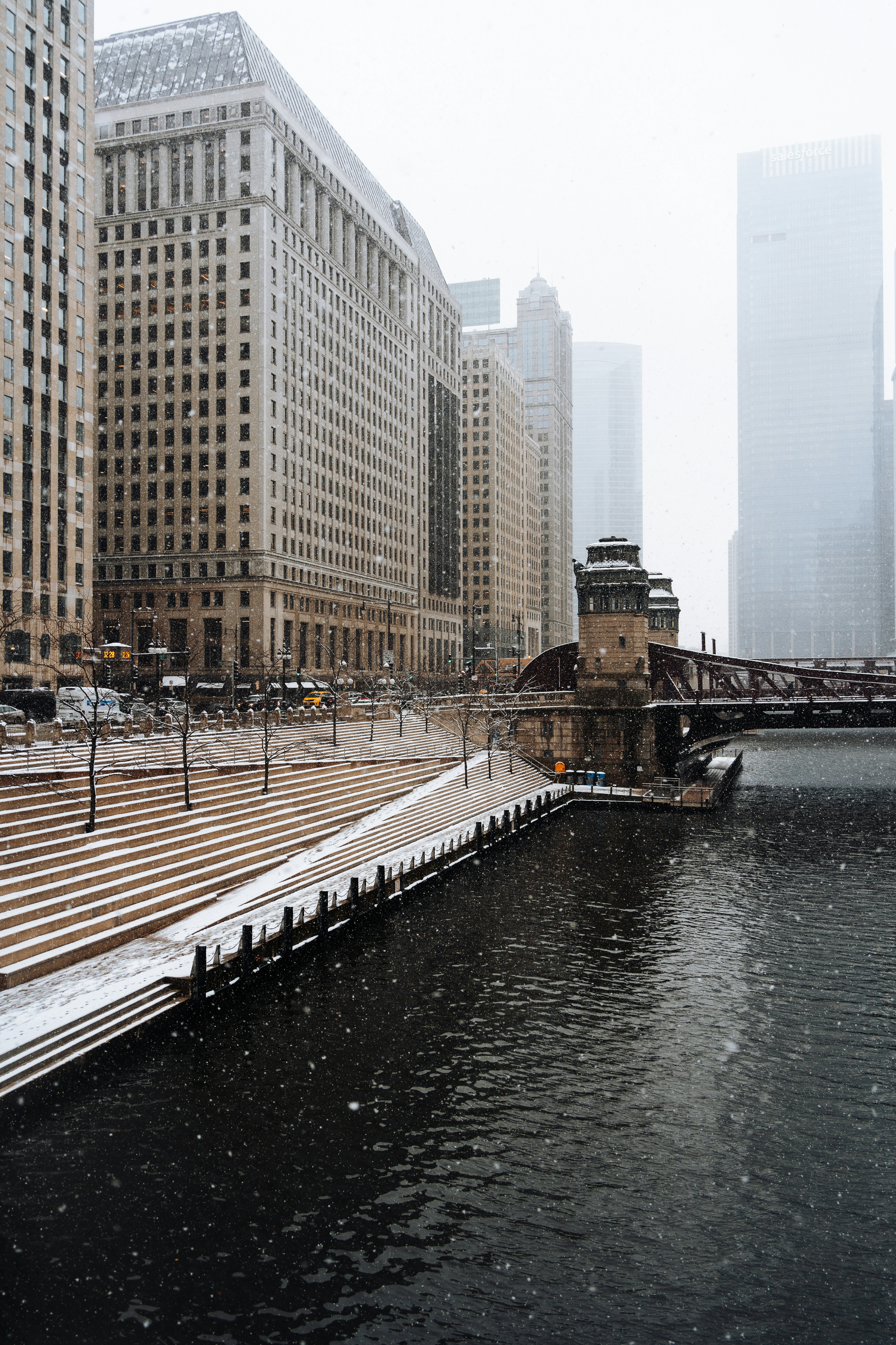 Snow-dusted riverbank flanked by towering city buildings under gentle snowfall.