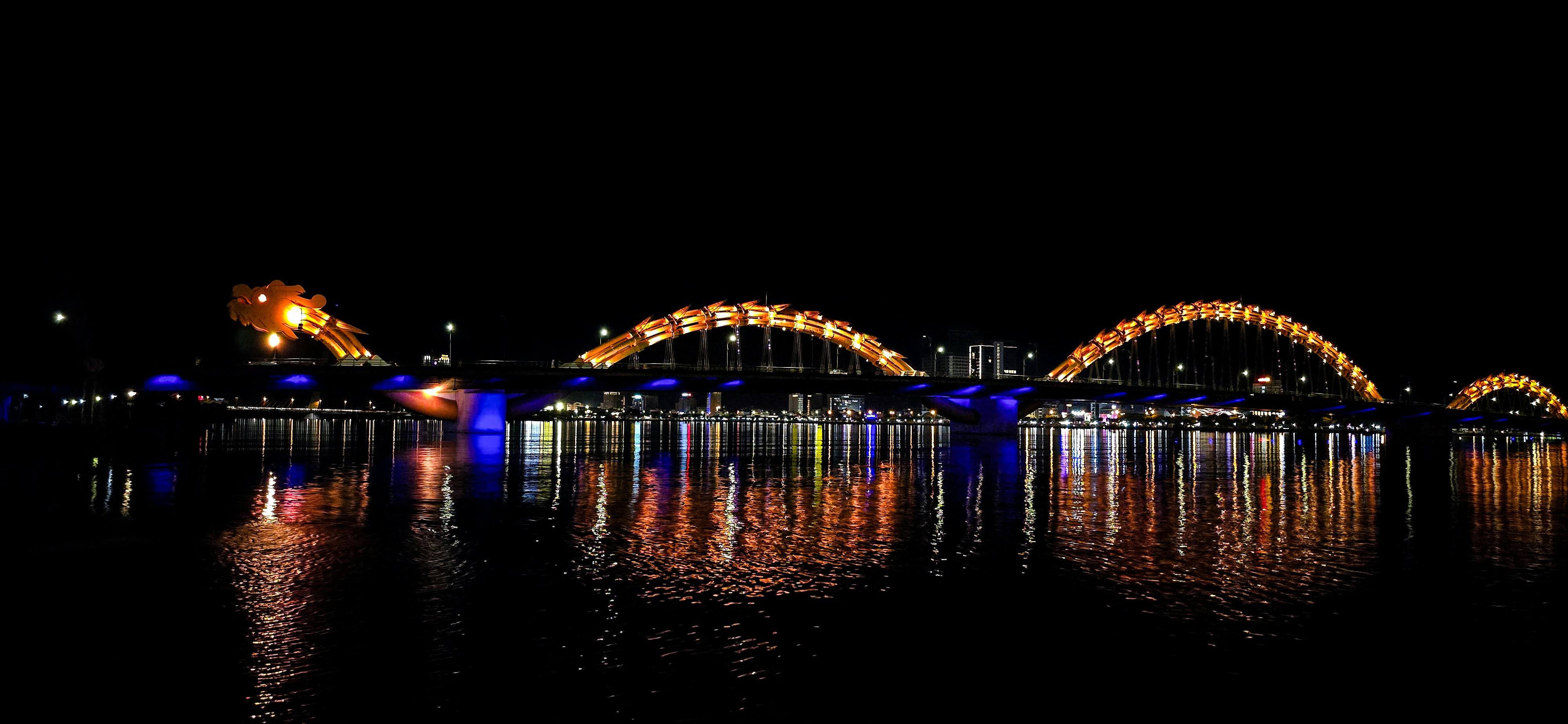 Golden dragon bridge illuminated at night, reflecting in the calm river below.