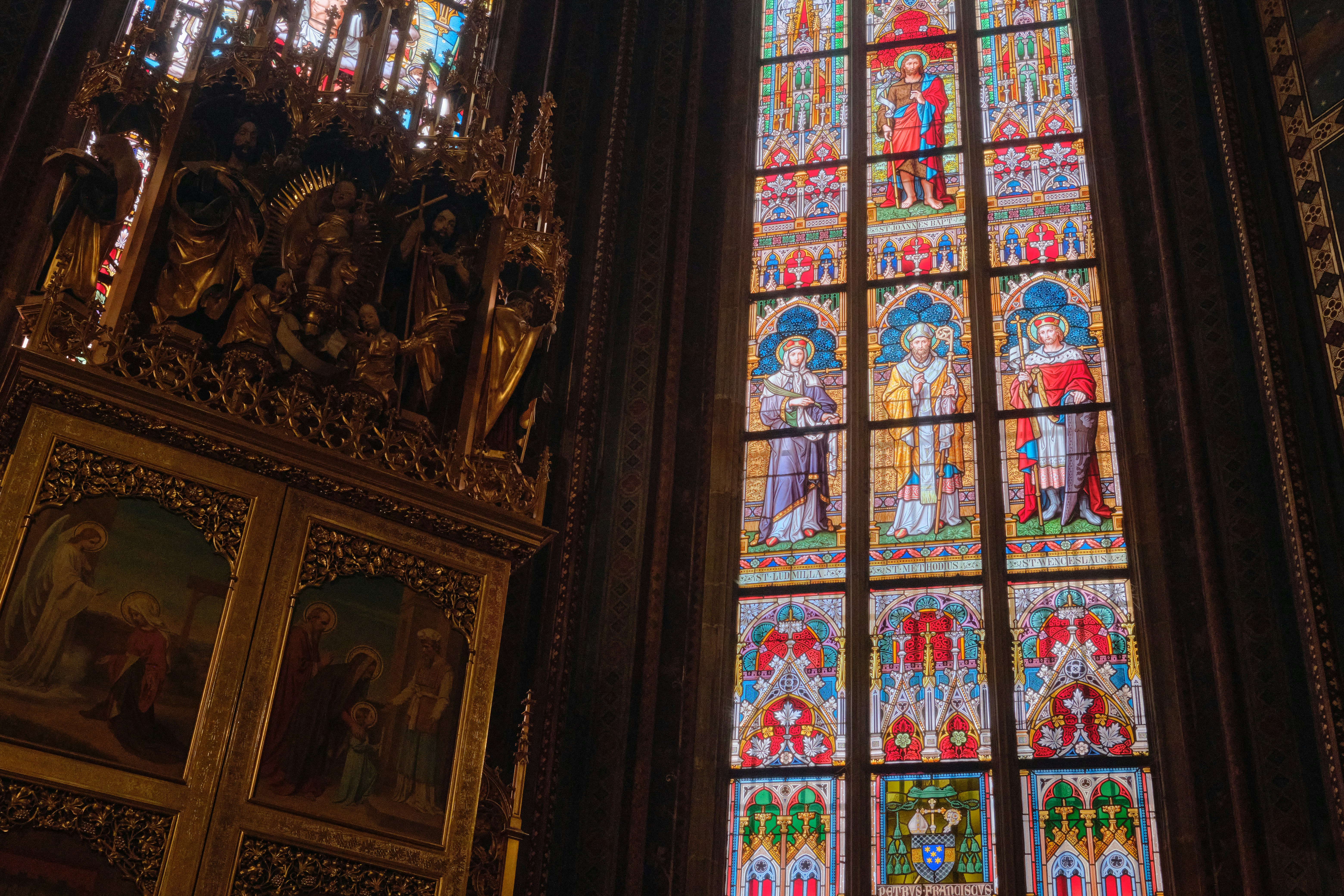 Intricate stained glass windows alongside ornate religious sculptures in a dimly lit cathedral.