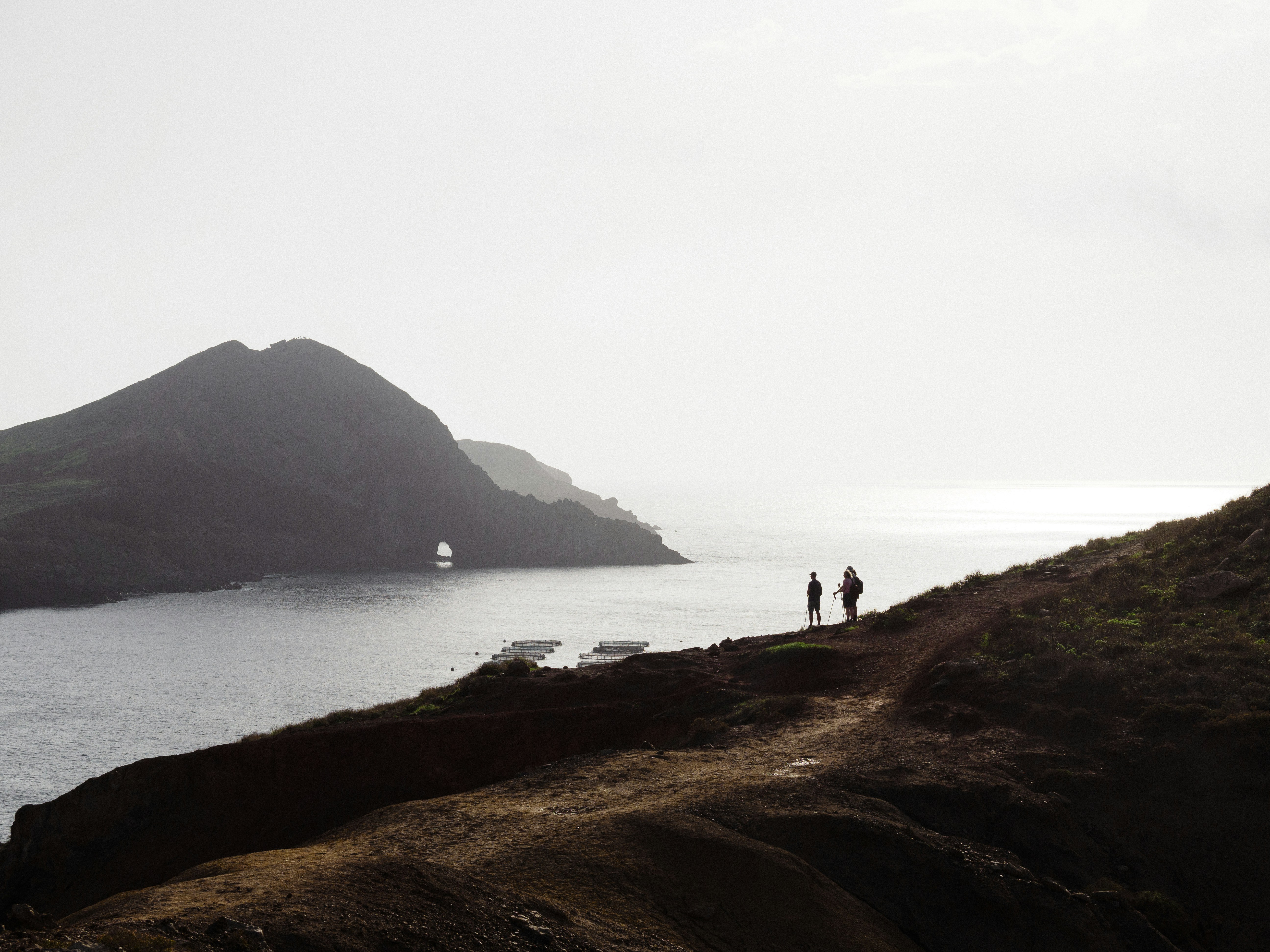 Two hikers on a rugged coastal path approaching a sea arch with a hazy horizon and distant light.