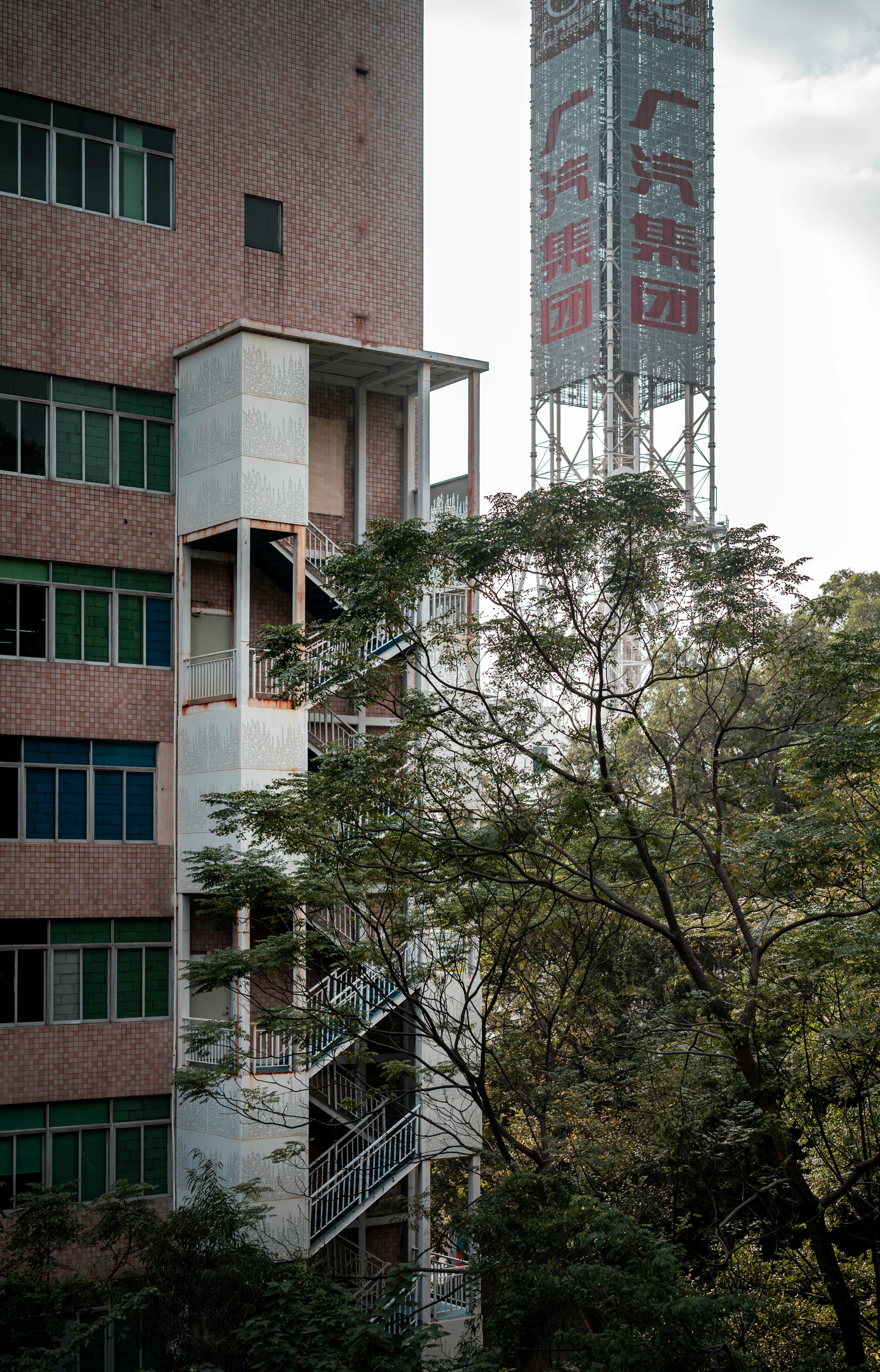 A multi-level external staircase ascends the side of a tall, brick-tiled building under an overcast sky. In the background, a large lattice advertising tower featuring red Chinese characters (广汽集团 - GAC Group) is visible. Green trees fill the foreground and mid-ground, partially veiling the lower structure and creating a contrast between the urban architecture and natural elements.